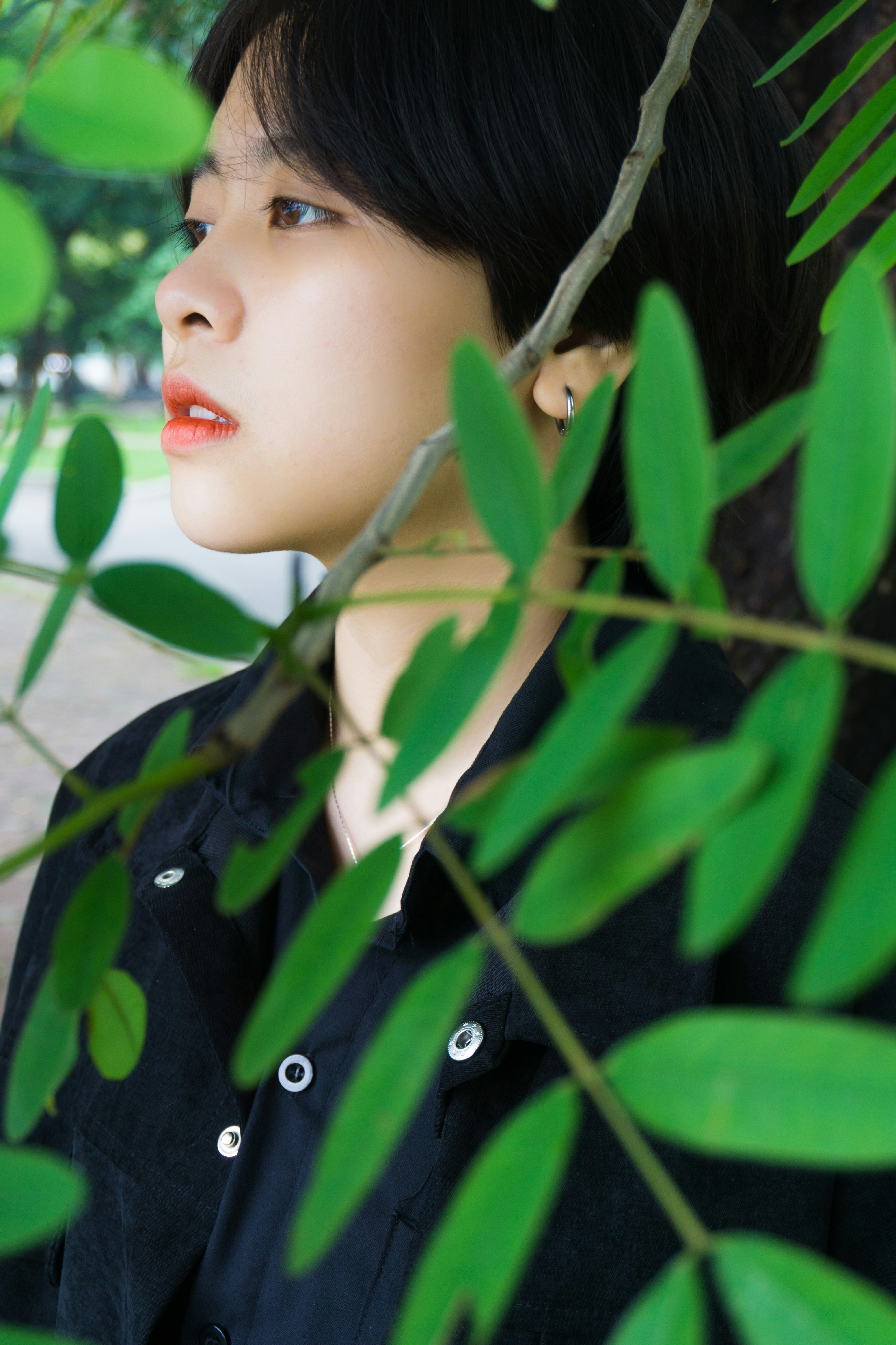 A young person gazes thoughtfully, partially obscured by vibrant green leaves, creating a natural frame around their profile. The scene highlights a harmonious blend of human presence and nature.