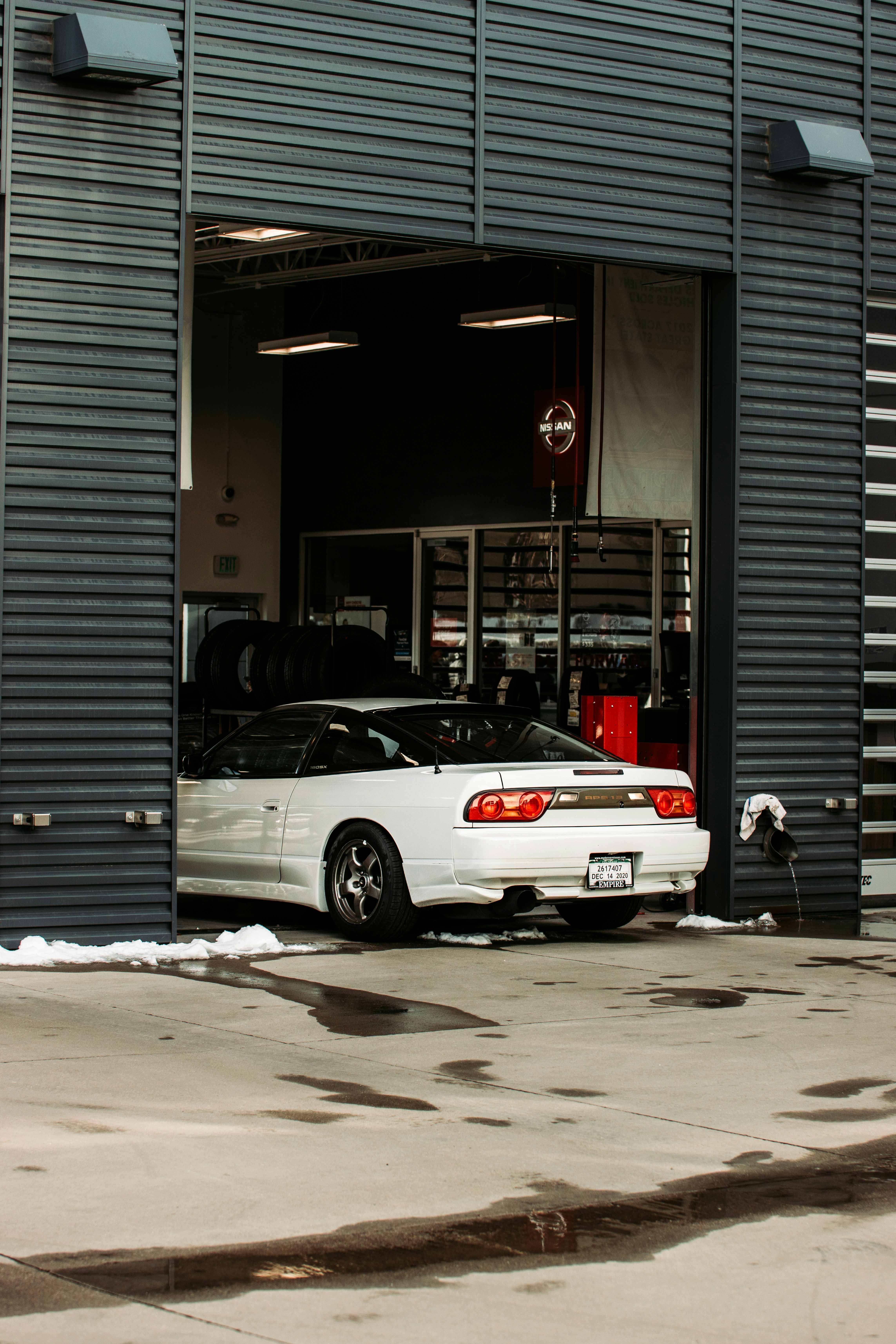 White sports car partially exiting a garage, framed by industrial architecture and snow remnants on the ground.