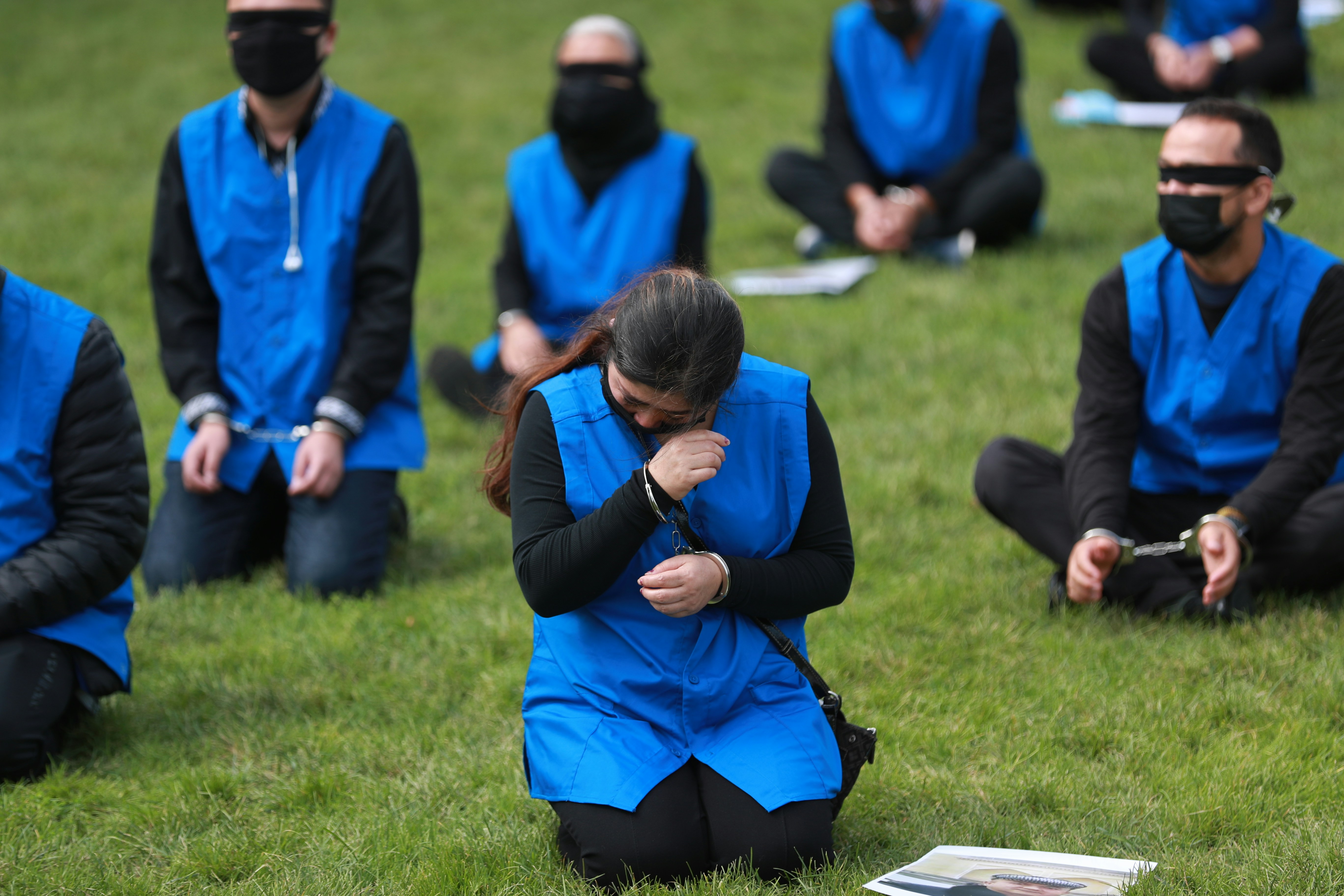 Participants in blue vests kneel on grass, some with masks, as they engage in a solemn gathering, expressing deep emotions during a moment of remembrance.
