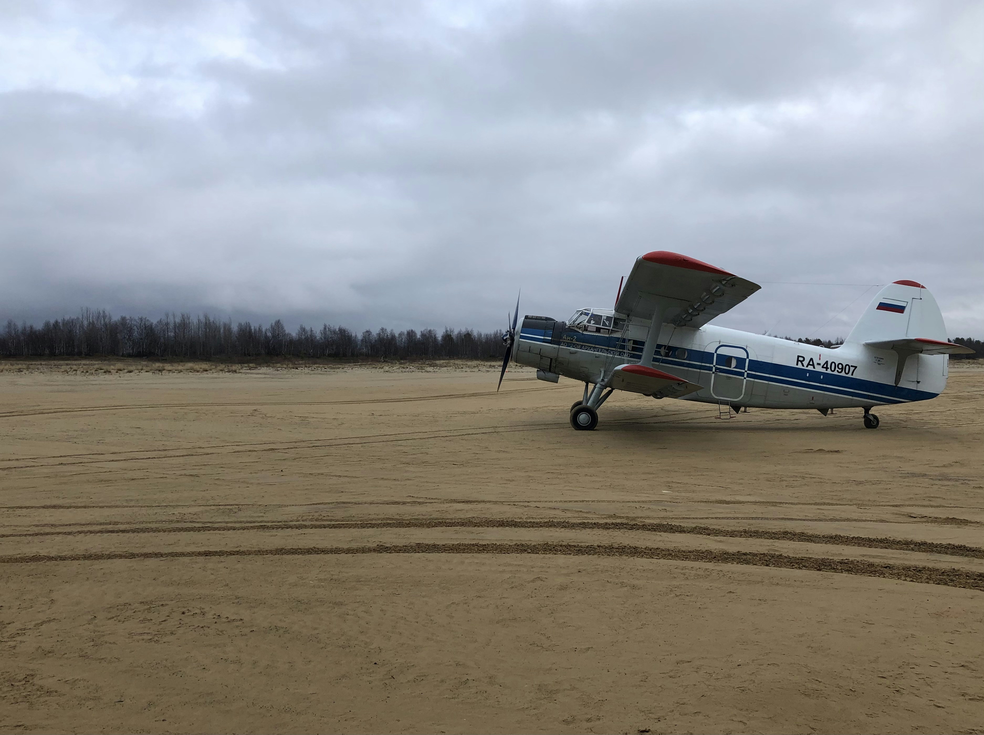 An Antonov An-2 biplane parked on a sandy landscape under a cloudy sky, showcasing its vintage design and rugged charm.