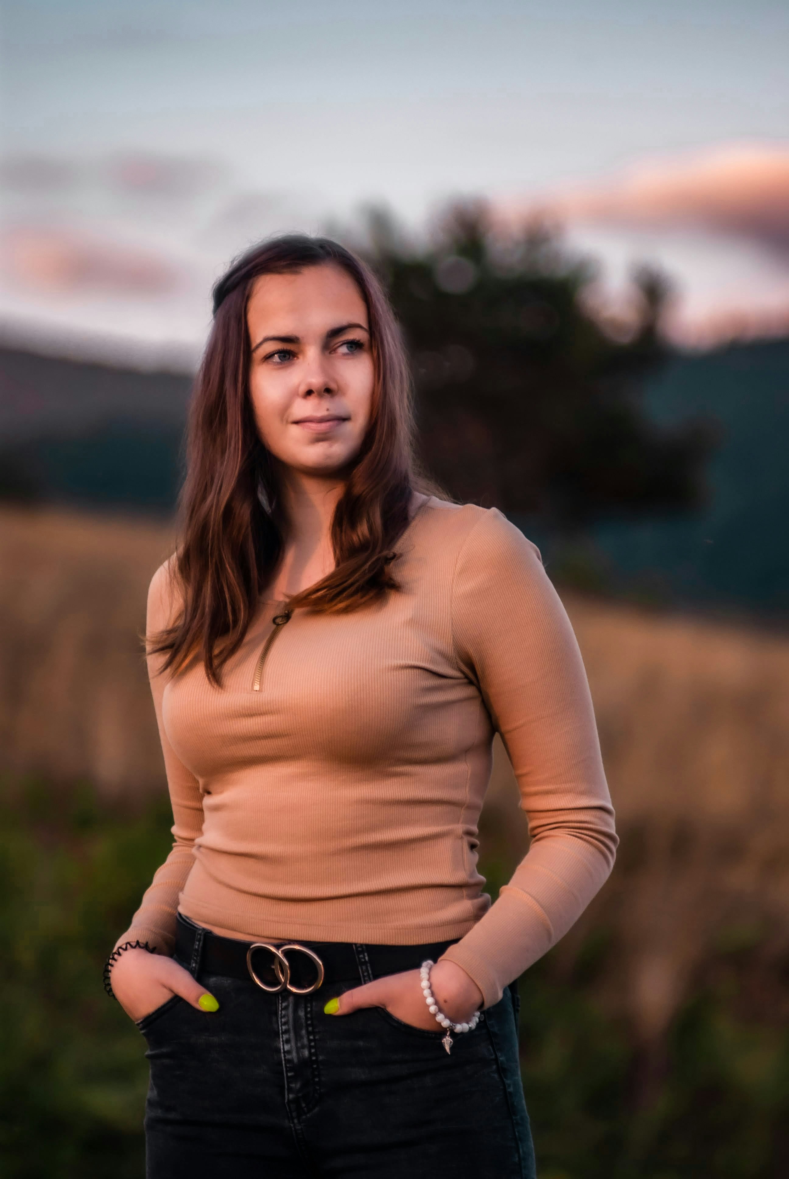 Young woman standing in a field at dusk, with a soft, blurred background of hills and trees. Her relaxed pose and thoughtful expression evoke a sense of calm.