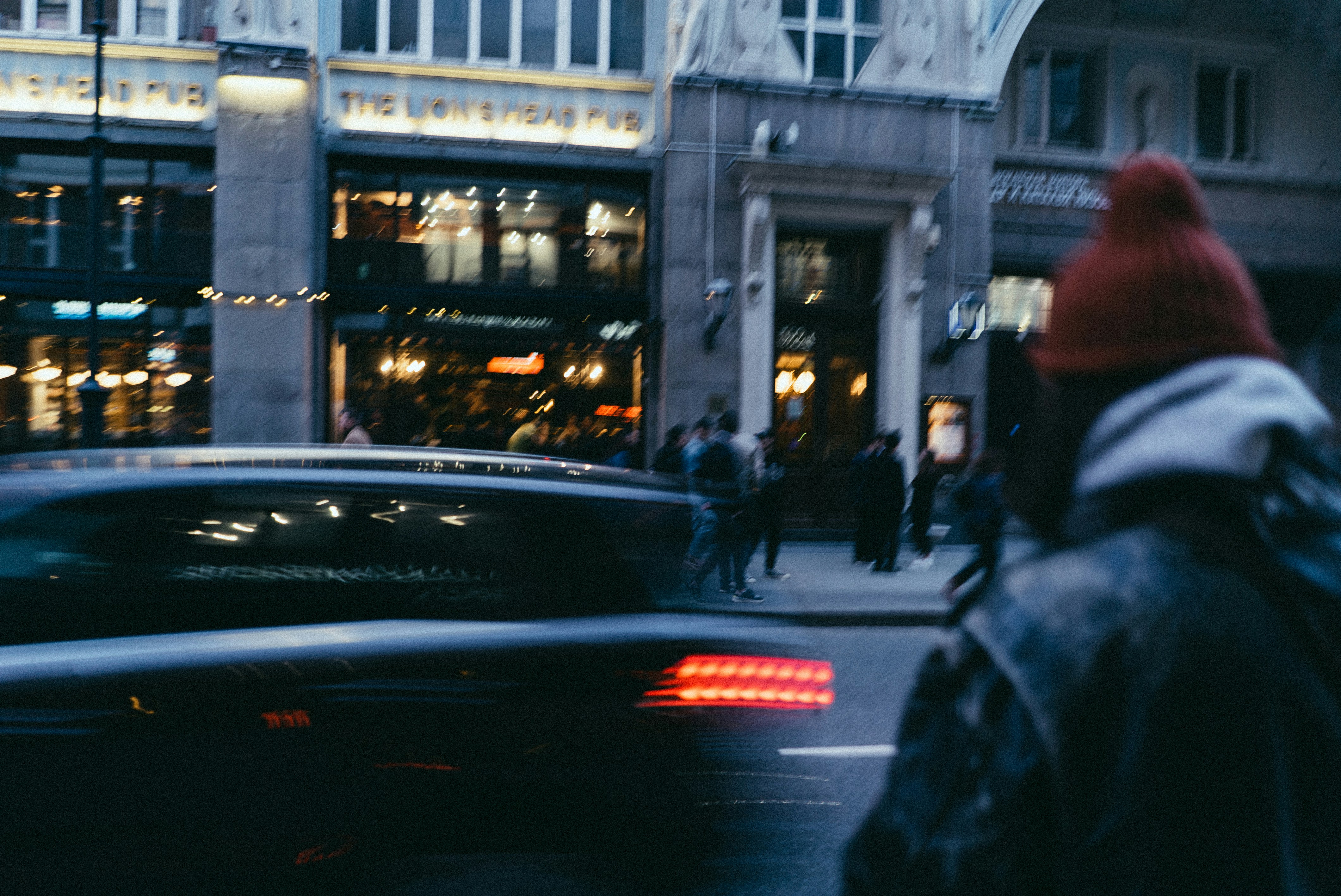 A figure in a red beanie stands still on a bustling street, while a blurred car speeds by, capturing the essence of urban life in motion.