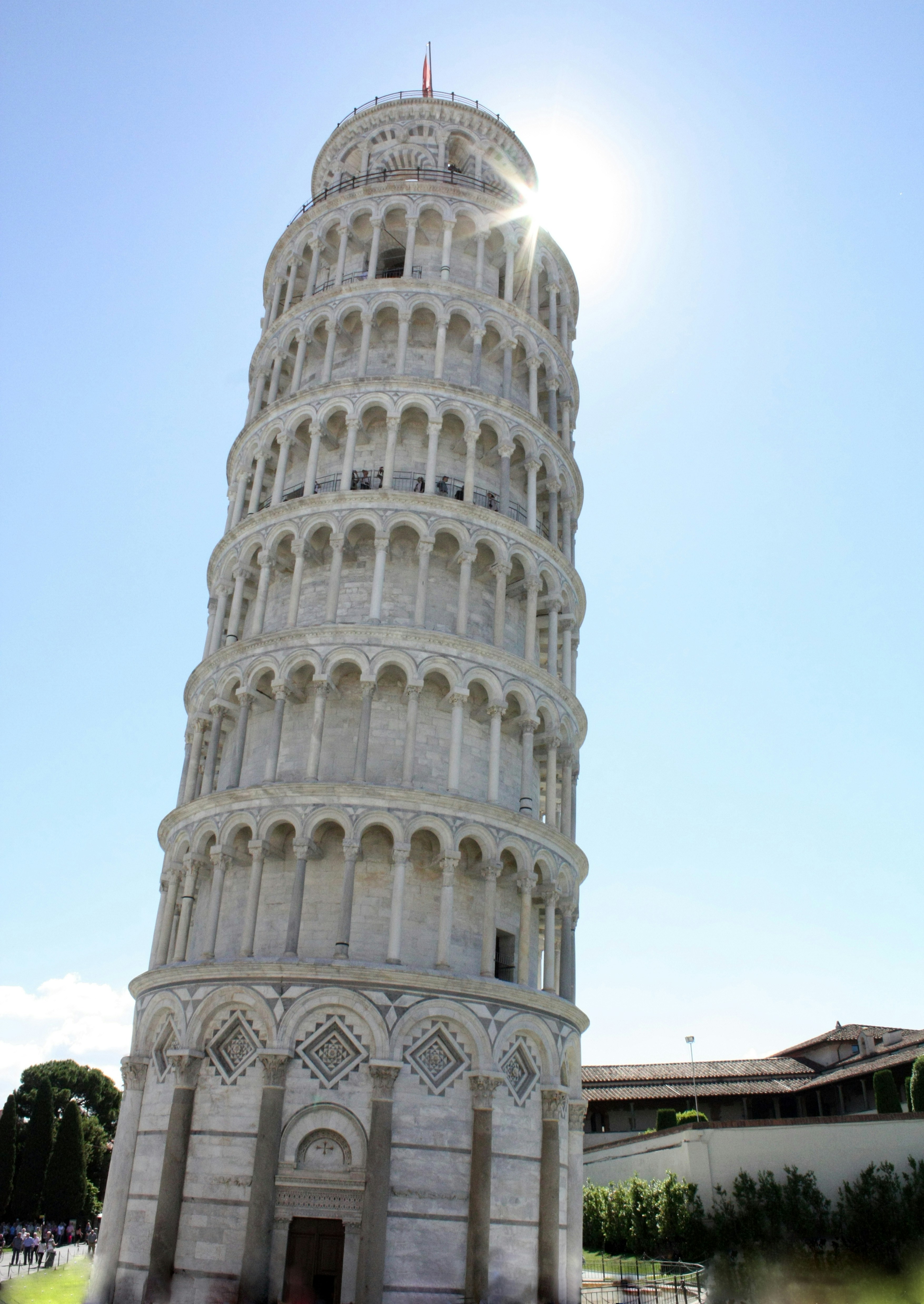 The Leaning Tower of Pisa stands tall against a clear blue sky, showcasing its iconic tilt and intricate architectural details. A hint of sunlight creates a halo effect around the top.