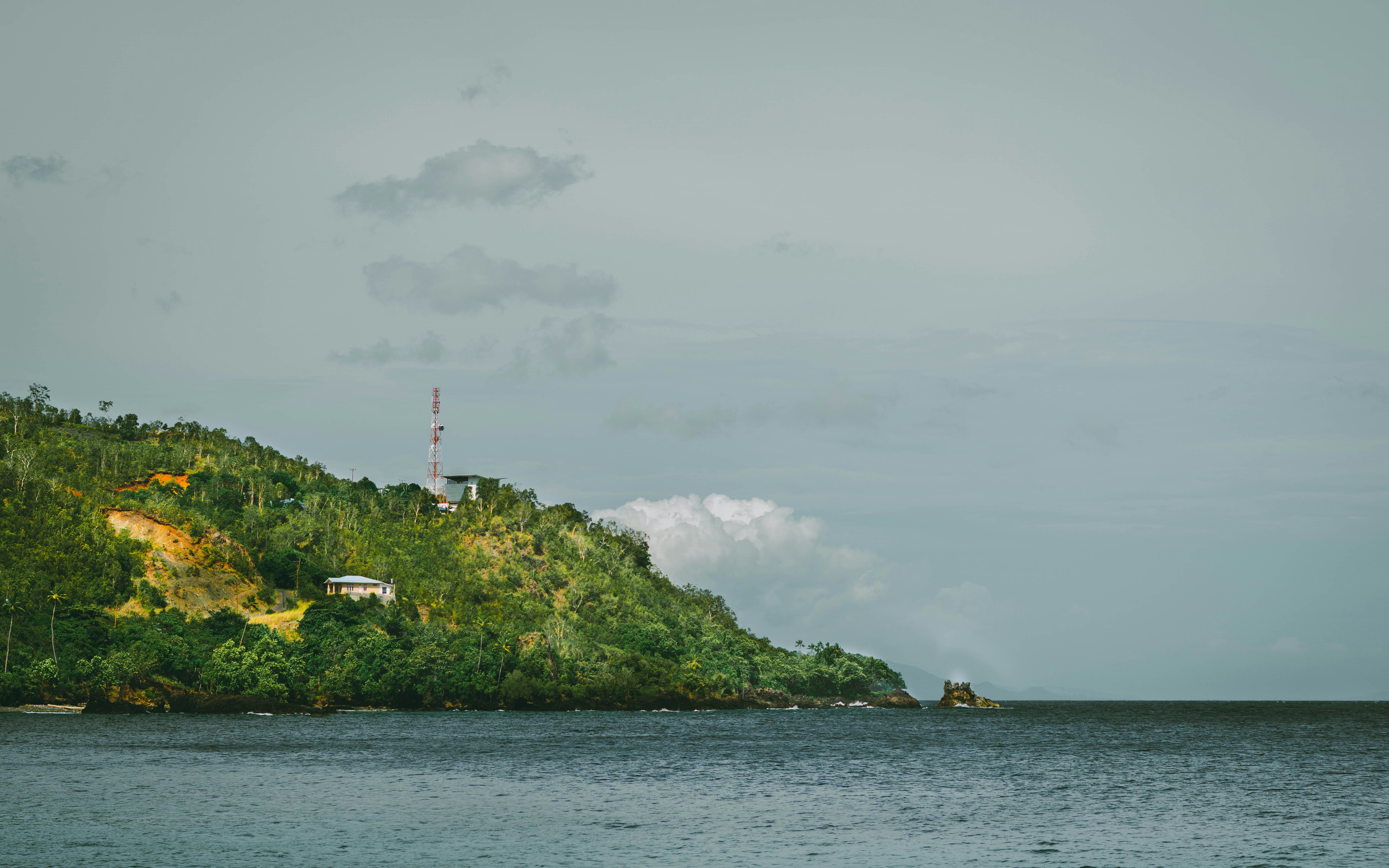 Lush green hillside rises above tranquil waters, featuring a communication tower and a solitary building. The scene captures the serene interaction between land and sea.