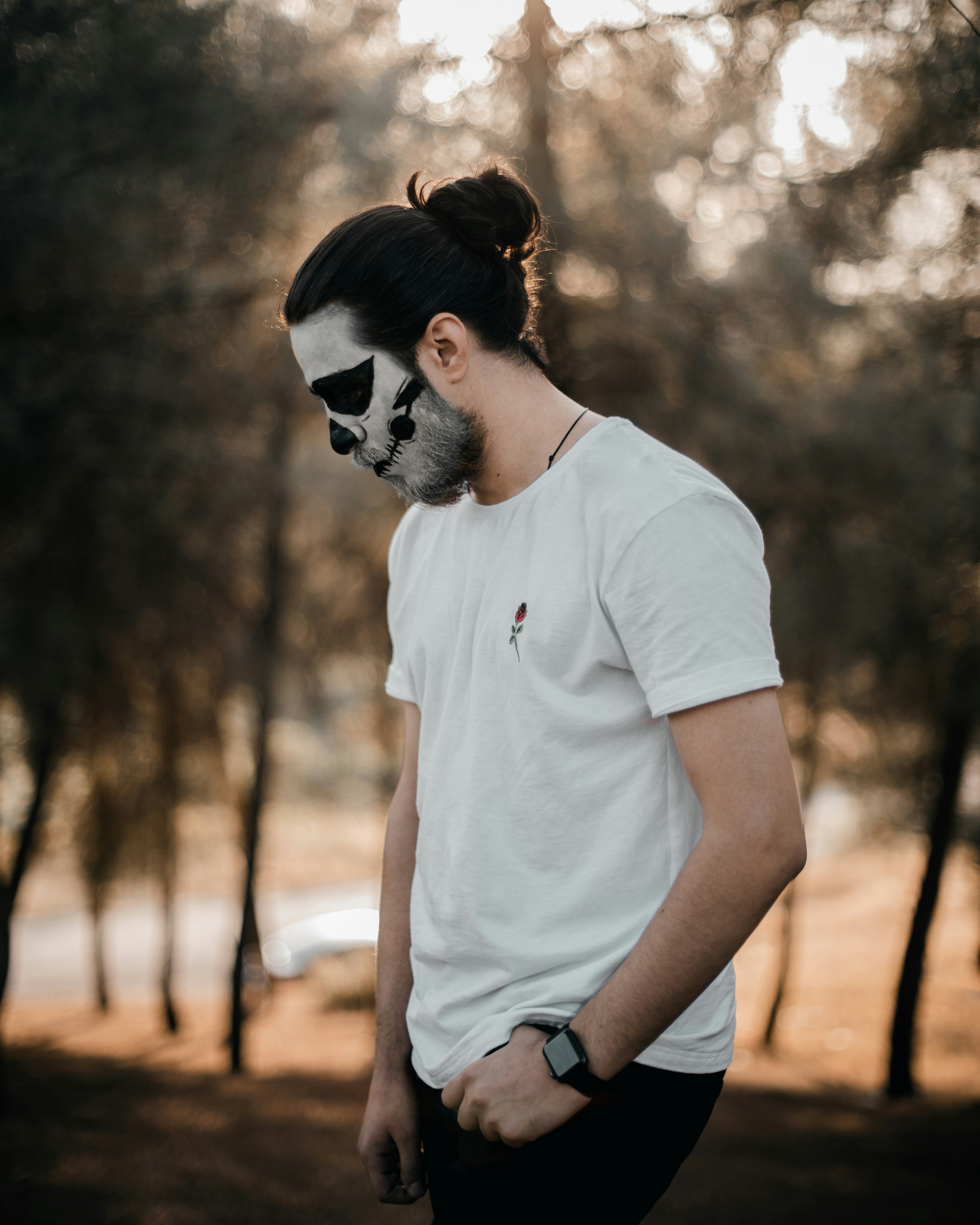 Young man with skull face paint stands contemplatively in a sunlit forest, embodying a blend of artistry and introspection.
