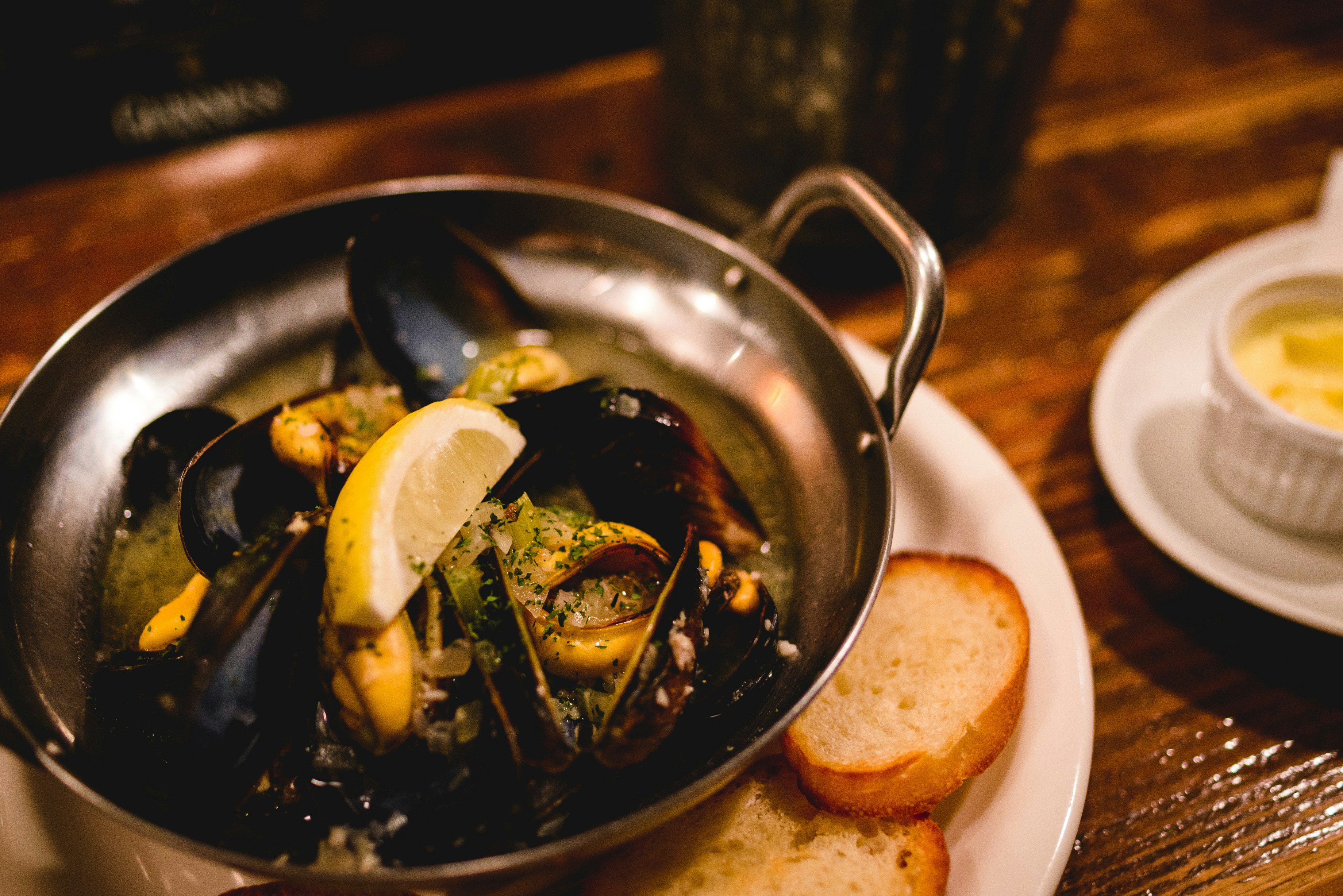 A serving of mussels garnished with lemon and herbs, accompanied by toasted bread and a side of butter. The dish is presented in a metal bowl on a rustic wooden table.