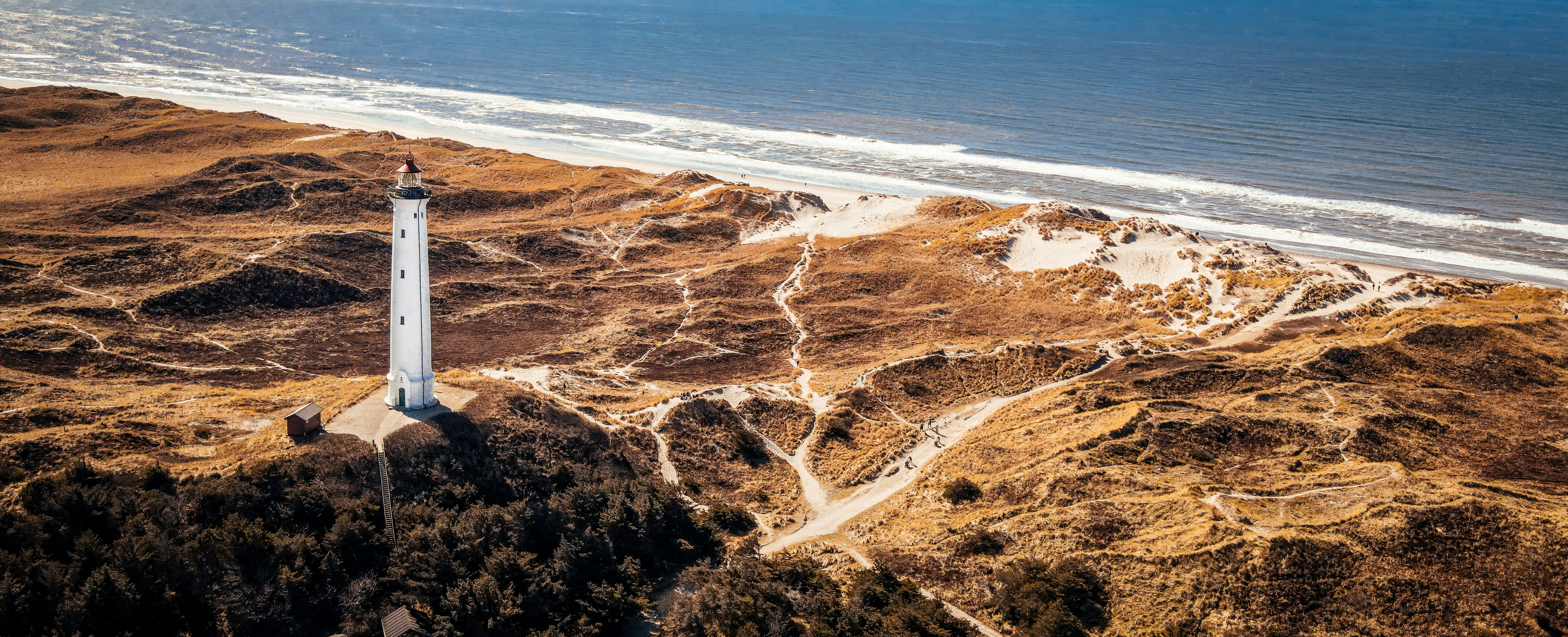 White lighthouse standing amidst rugged golden dunes beside a vast ocean expanse.