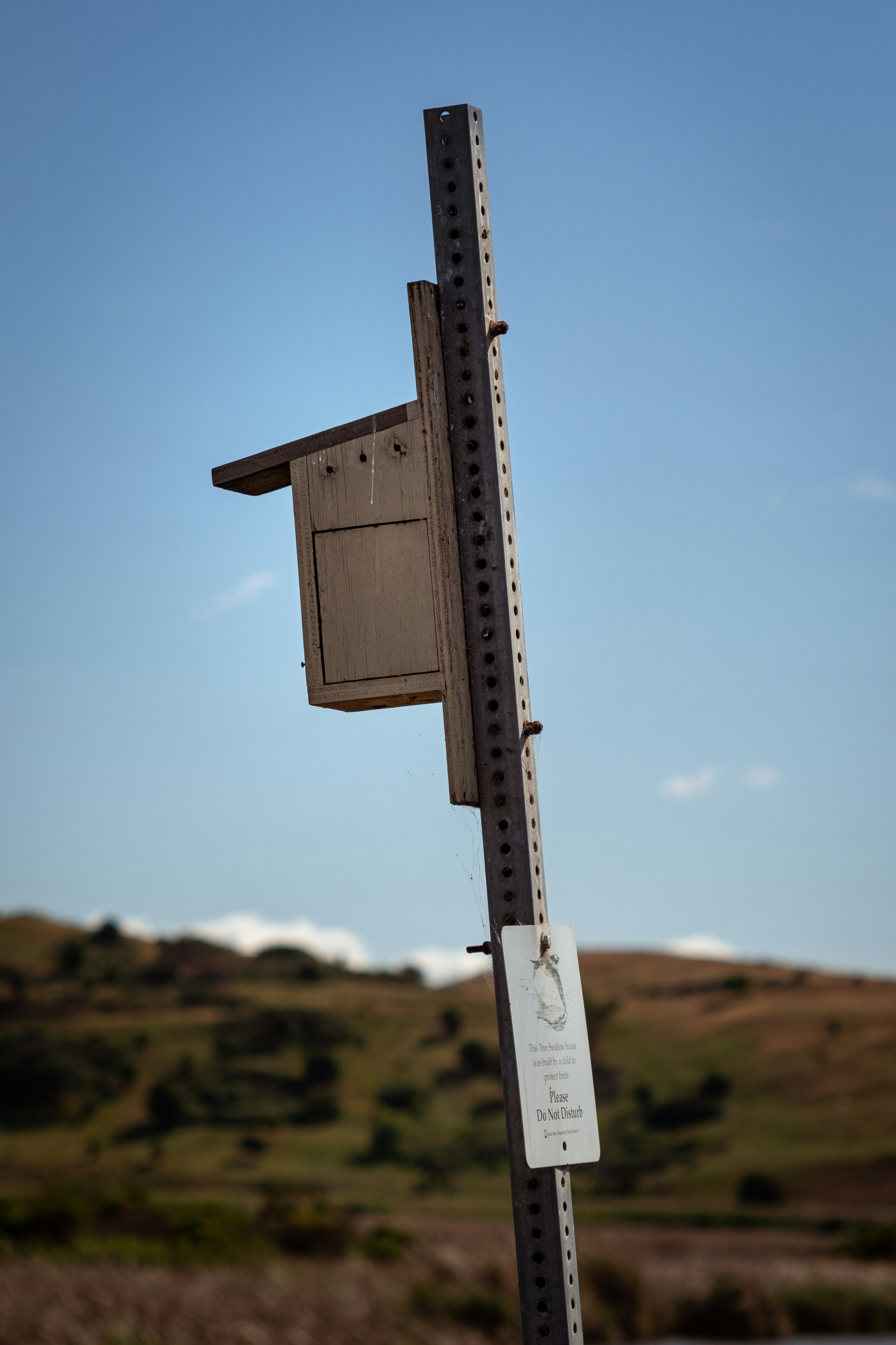 A weathered birdhouse mounted on a post, accompanied by a sign, set against a backdrop of rolling hills under a clear blue sky.