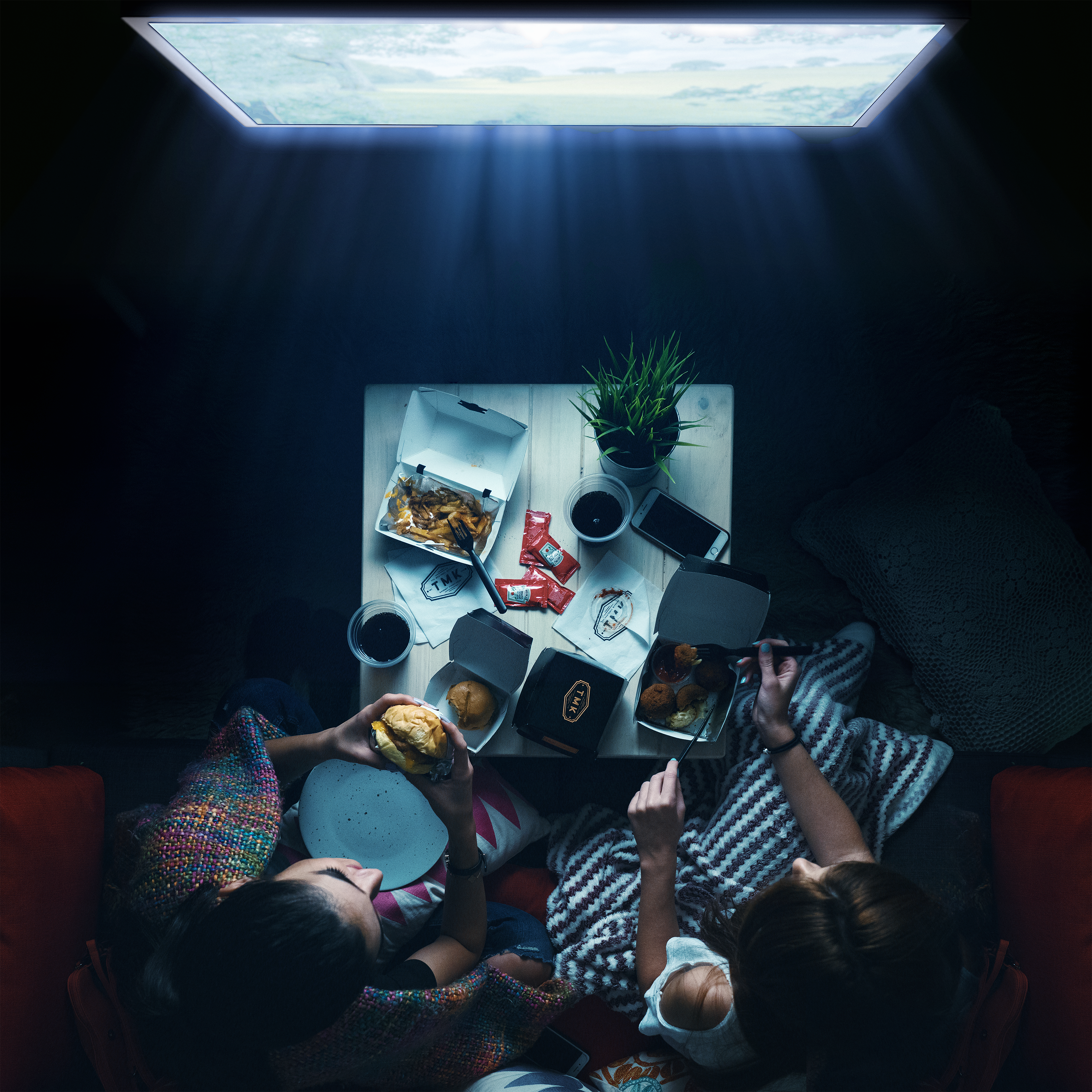 Two friends enjoying a meal at a table illuminated by soft rays of light streaming from a skylight. Takeout boxes and coffee cups create a cozy dining experience.