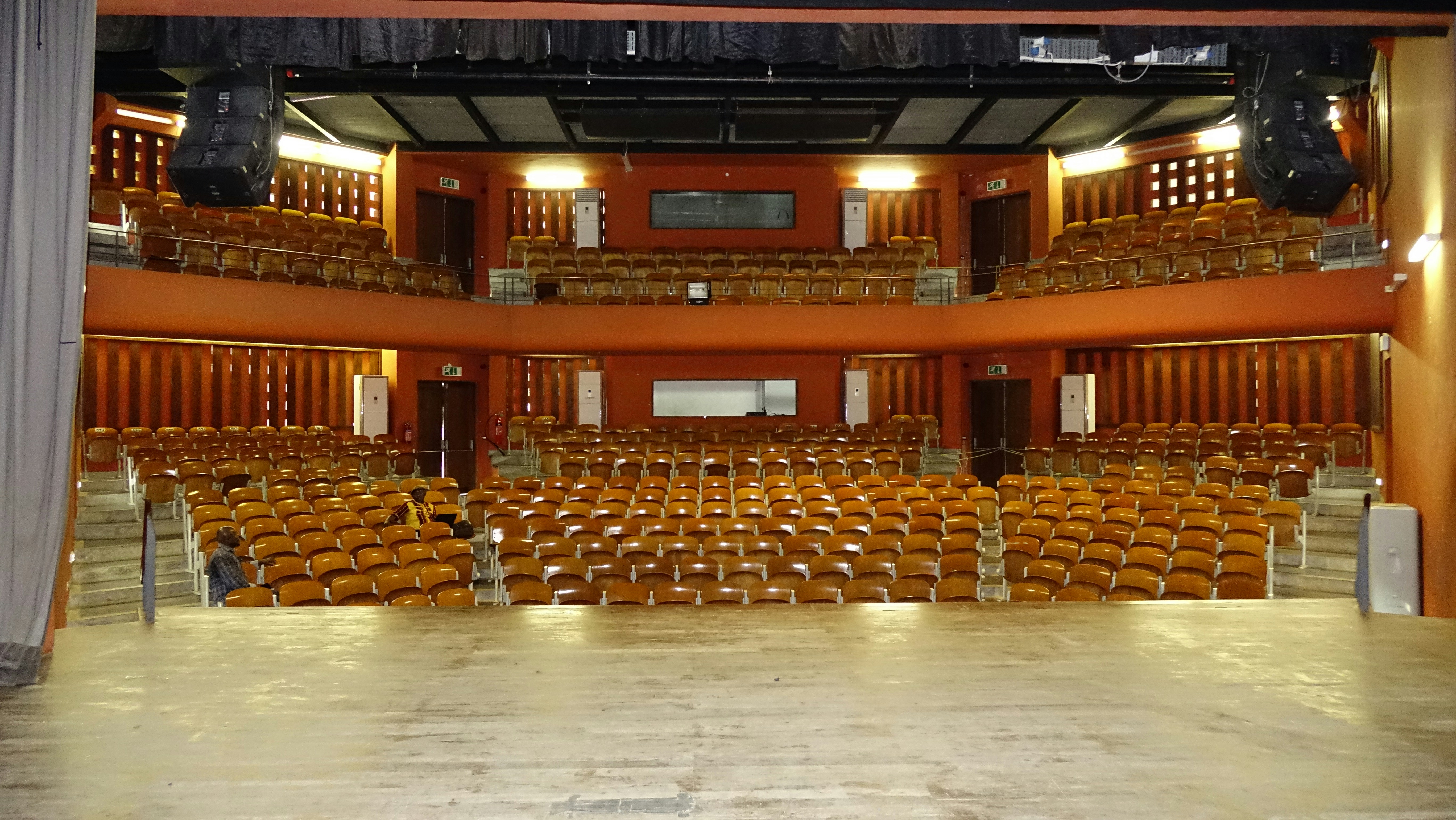 An empty theater photograph with warm amber lighting and rows of brown seats facing a stage, viewed from the proscenium area.