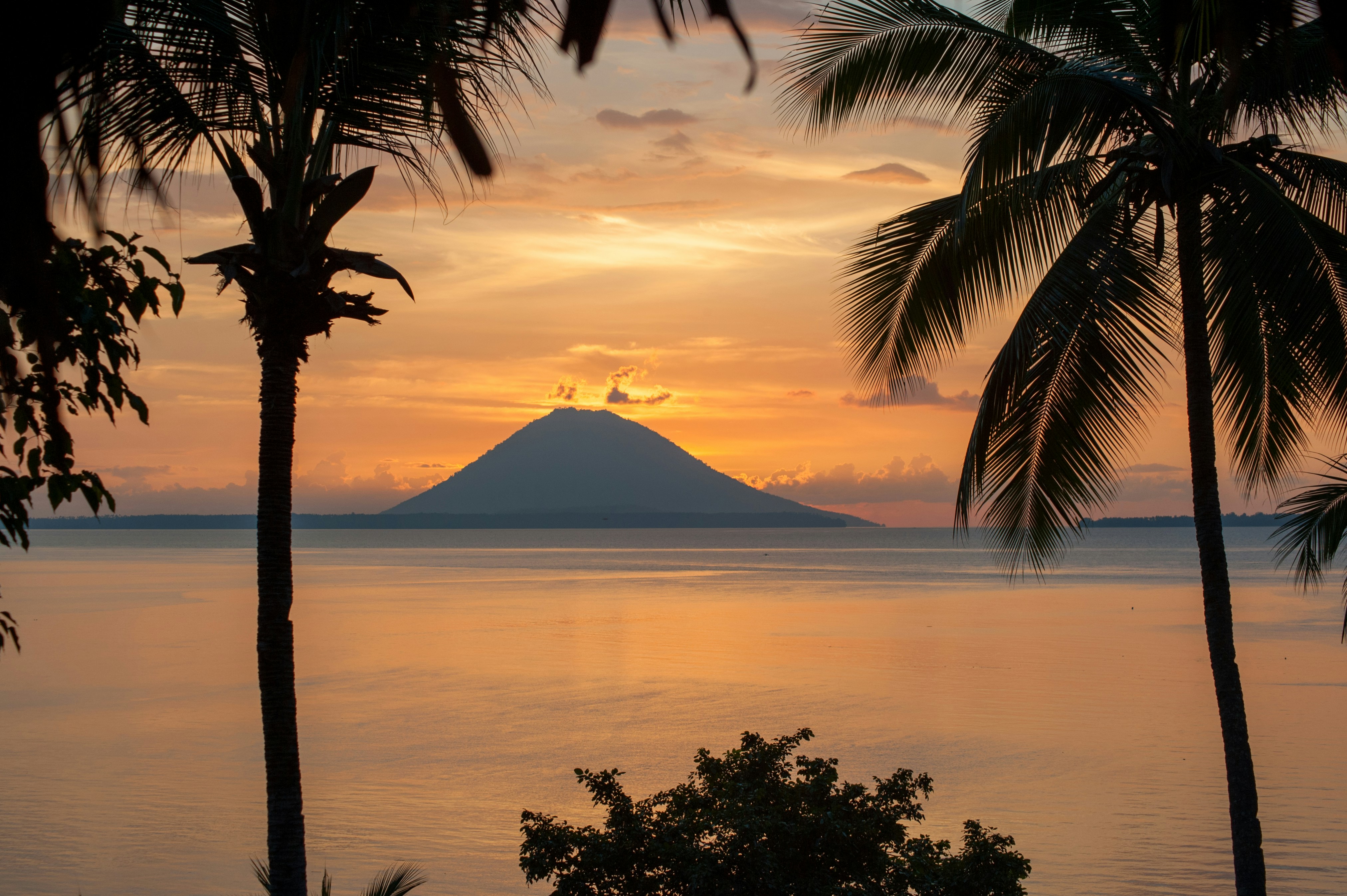Tropical sunset framing a distant volcano with palm trees and calm waters.