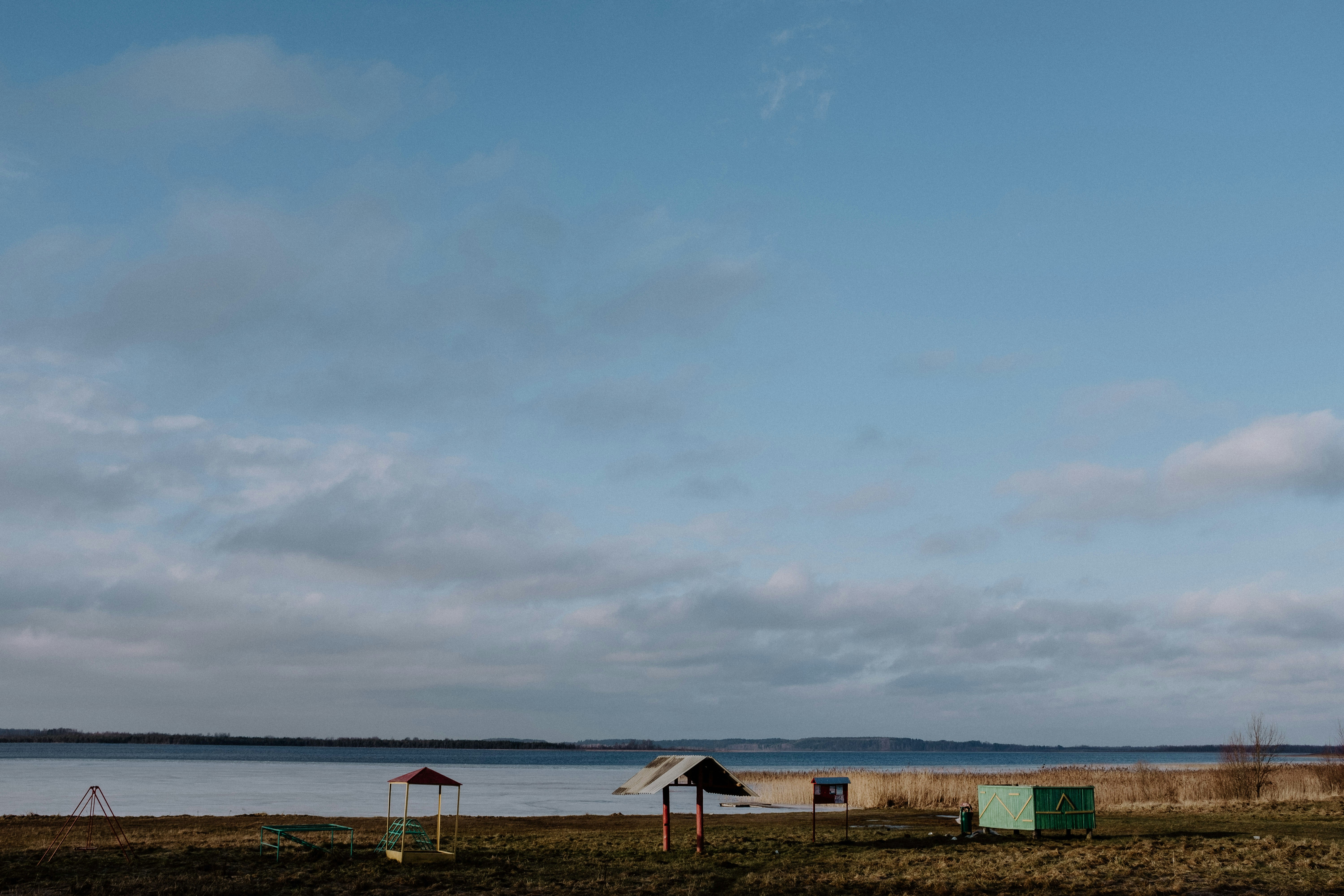 Open landscape with a calm lake, scattered clouds, and small structures on grassy shore.