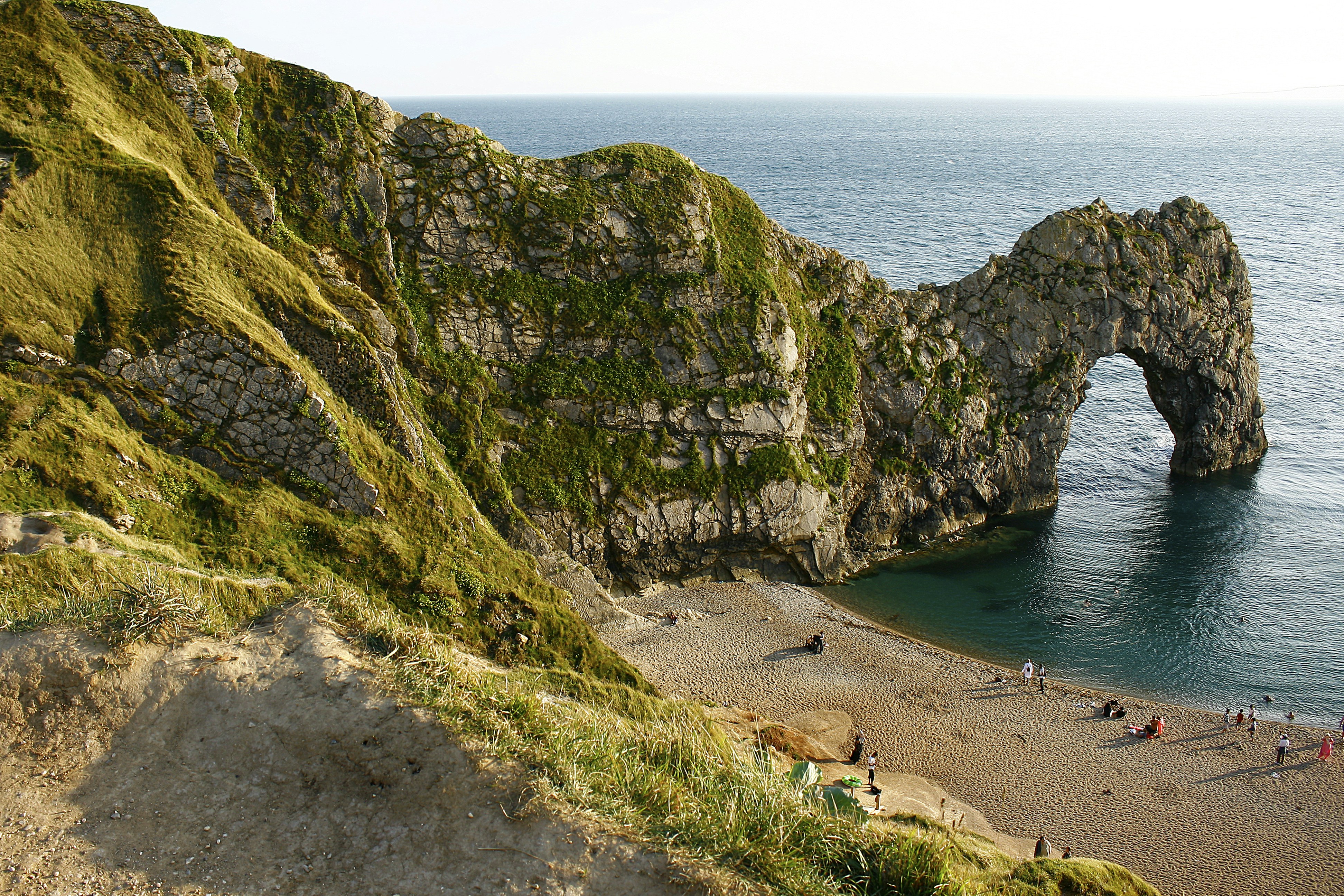 Dramatic limestone arch rising from the sea, surrounded by lush greenery and sandy beach, with visitors enjoying the shoreline.