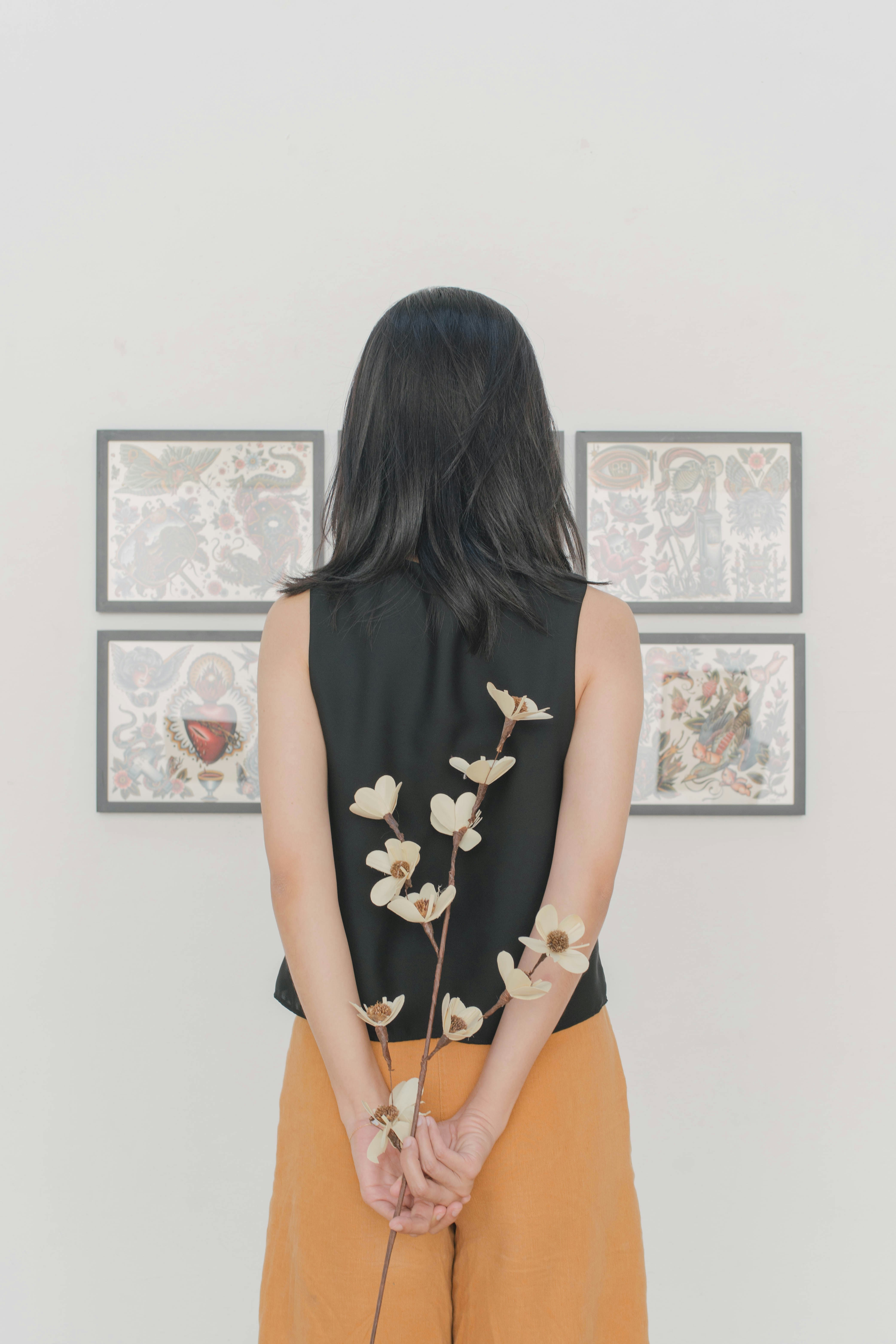 A woman stands with her back to the viewer, holding a delicate branch of flowers behind her while gazing at framed artworks on the wall.