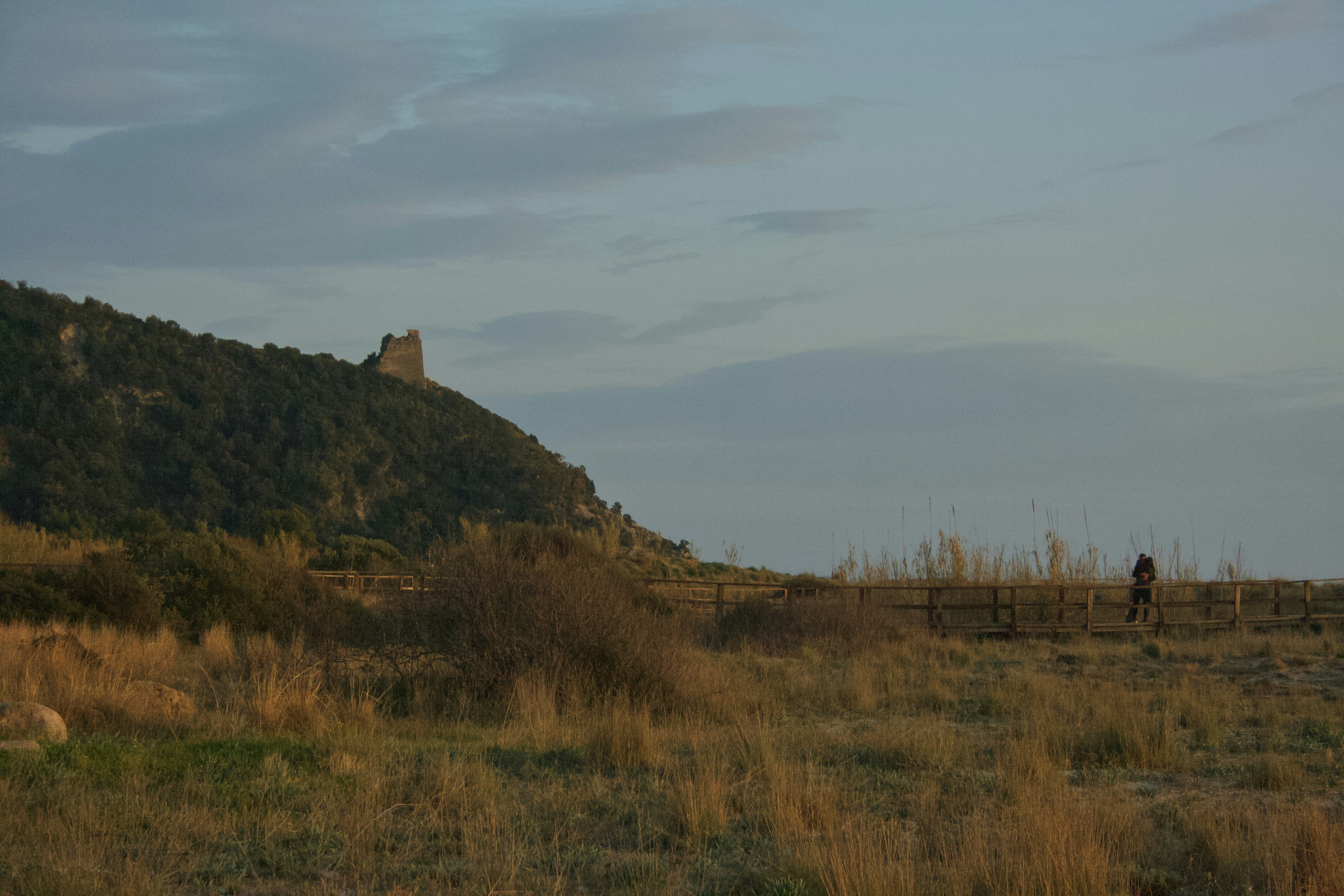 Ruins of a stone tower rise above a grassy landscape under a soft evening sky, with a solitary figure walking along a wooden path.