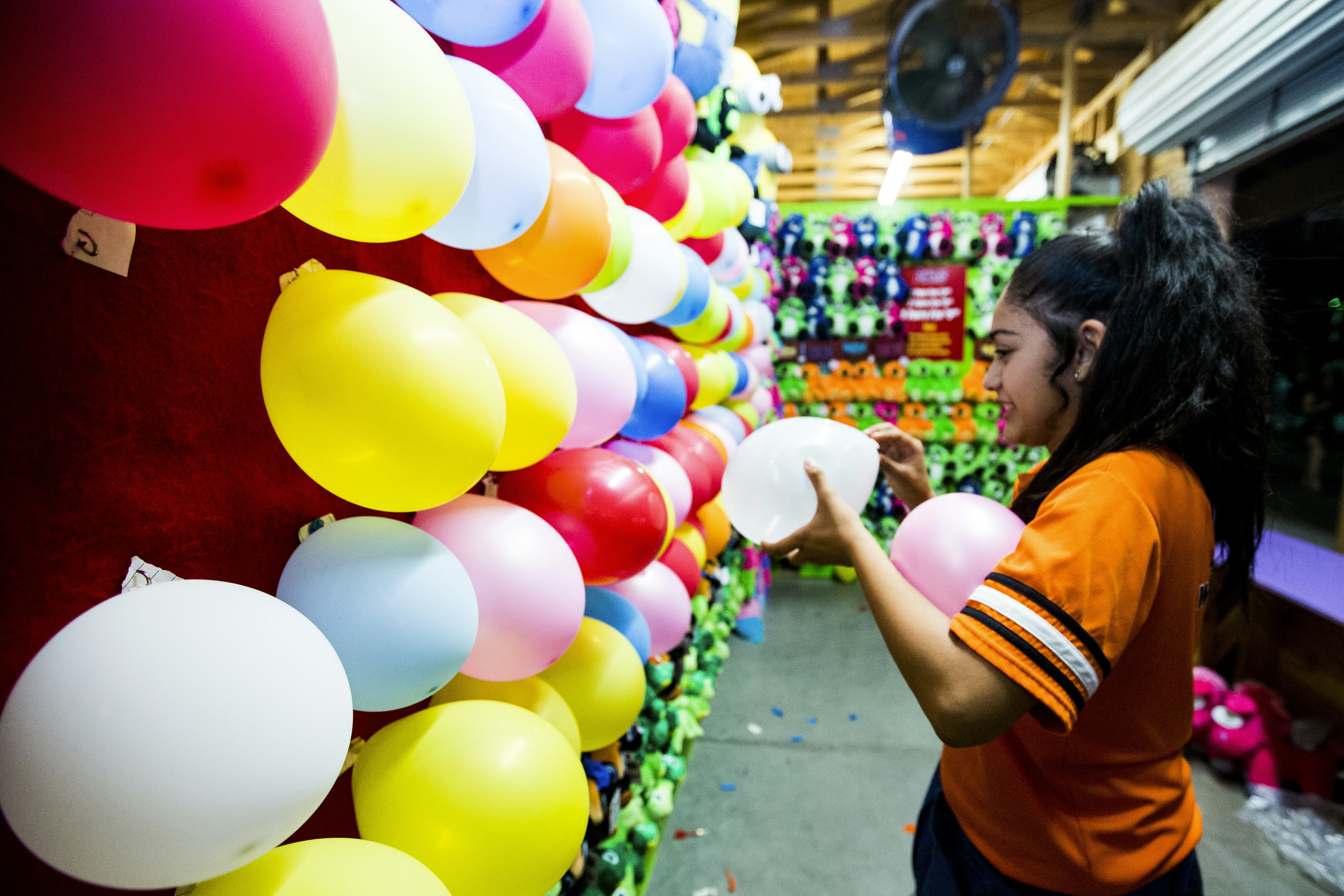 A young woman in an orange shirt selects a balloon from a vibrant wall of colorful balloons at a carnival game booth.