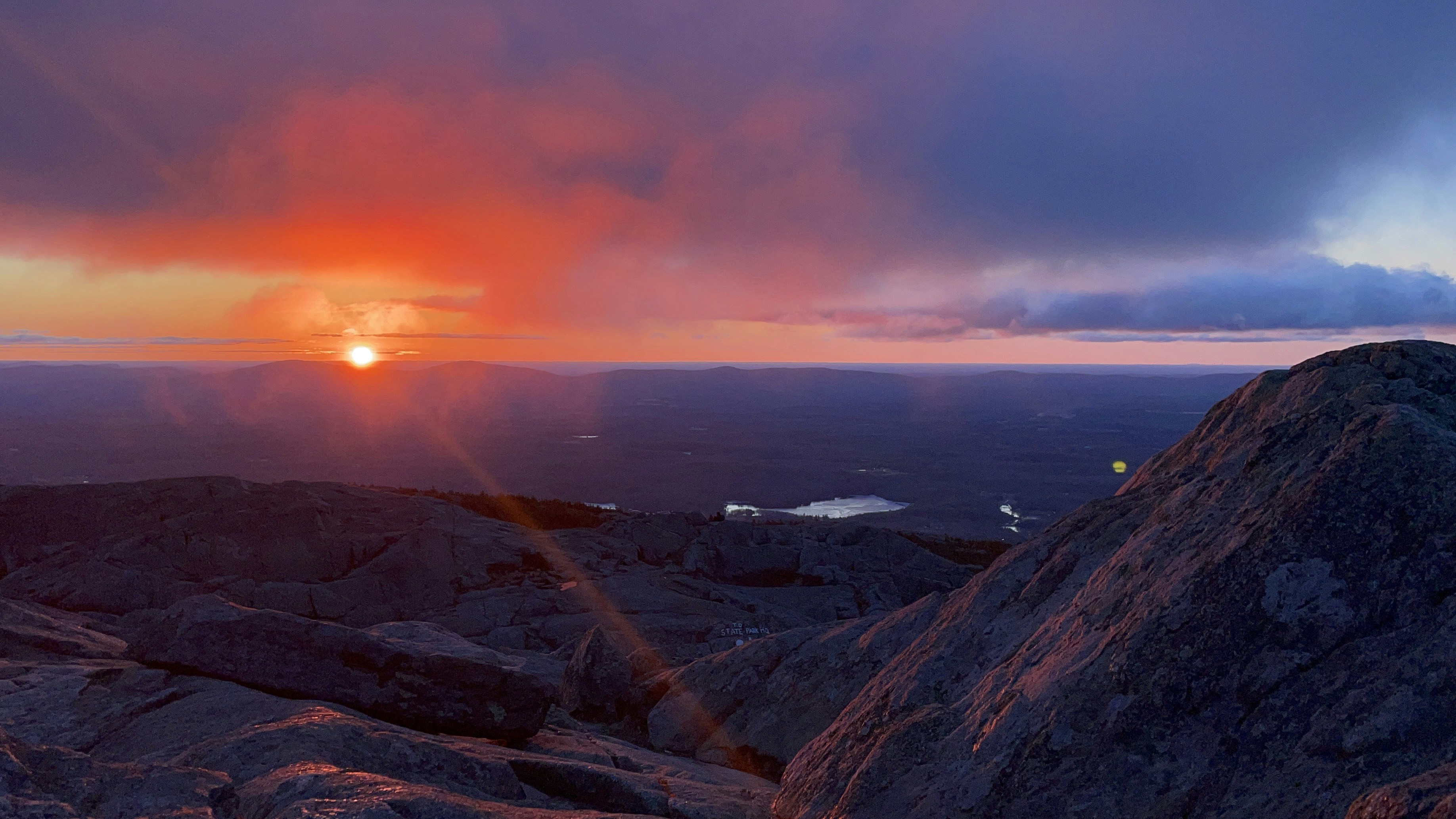 Vibrant sunset casting warm hues over rugged mountain terrain, with a prominent rock formation in the foreground.