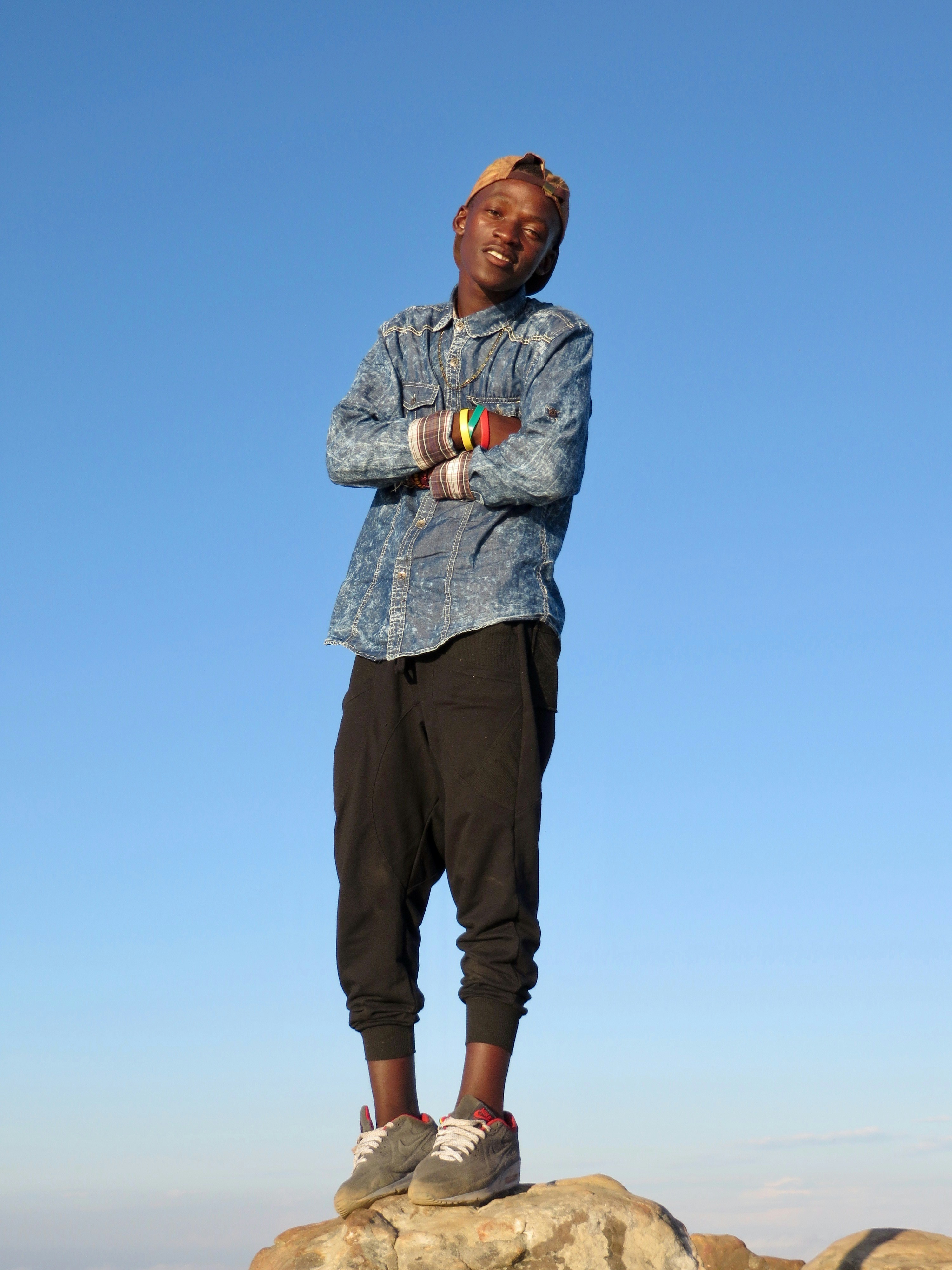 Young person standing confidently atop a rocky outcrop against a clear blue sky, arms crossed with a relaxed posture.