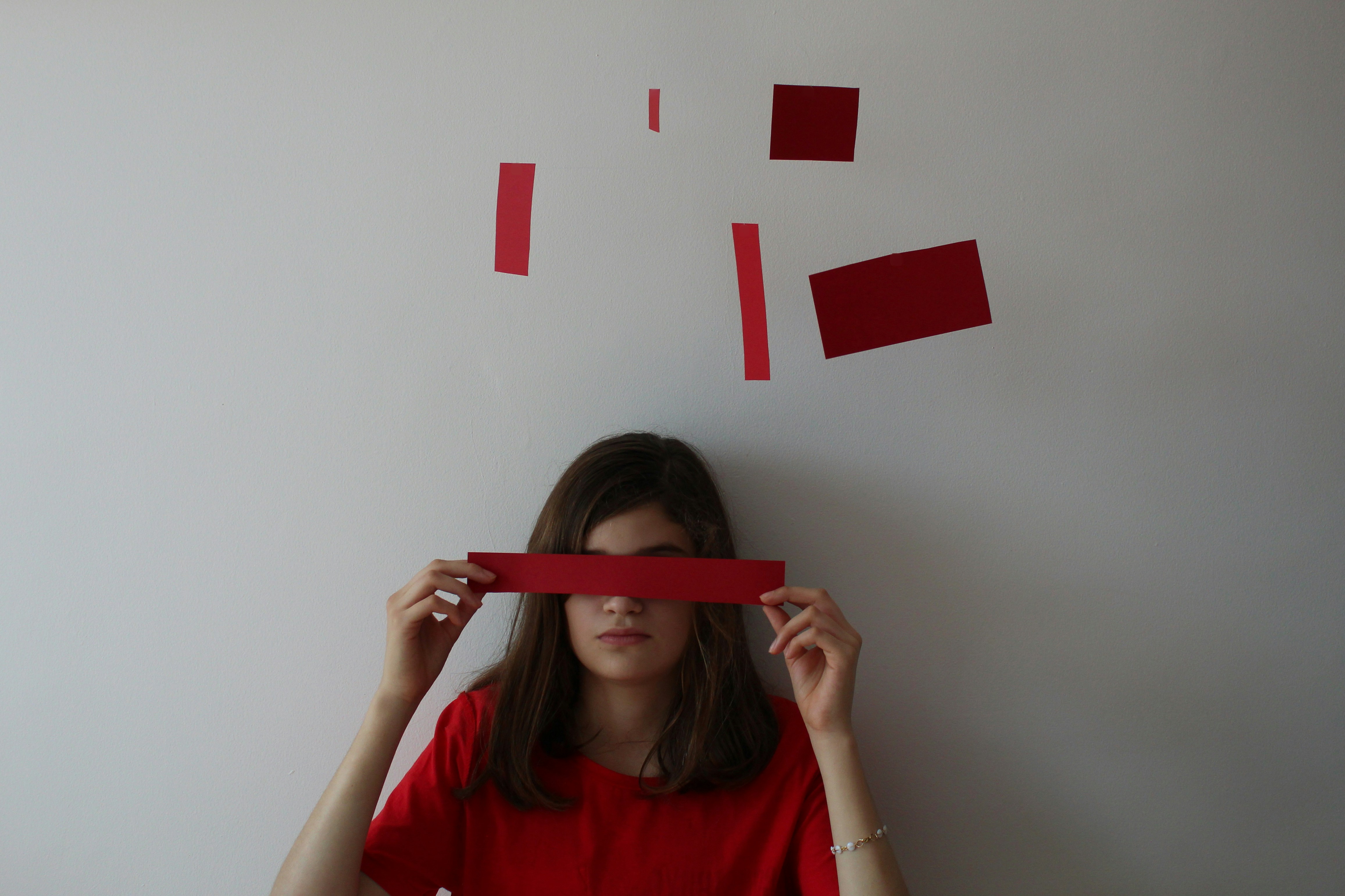 Person in red shirt holding a red strip over their eyes with red rectangles floating above against a plain background.