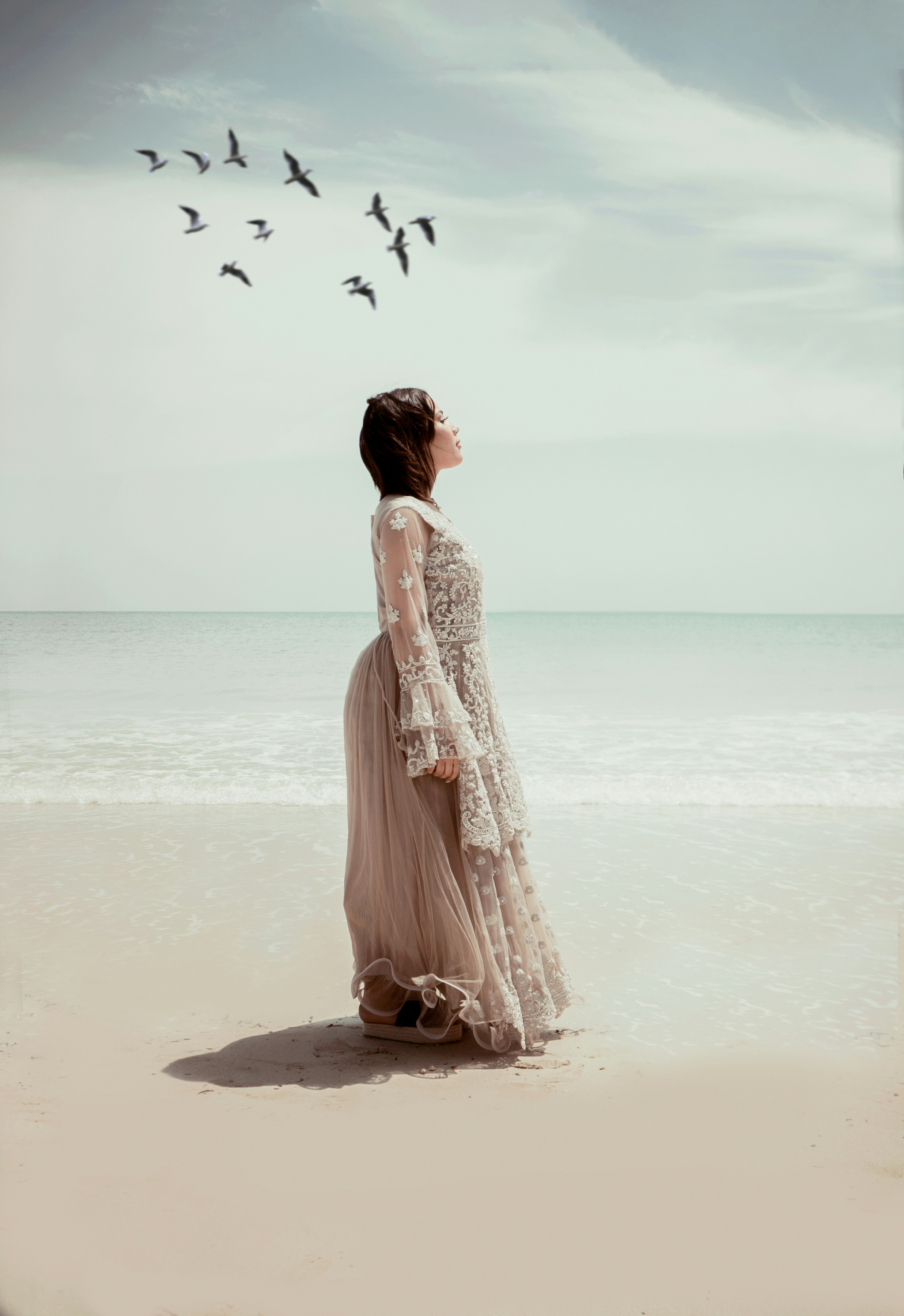 A woman in an elegant dress stands on the shore, gazing skyward as birds fly overhead. The serene beach setting enhances the moment's tranquility.