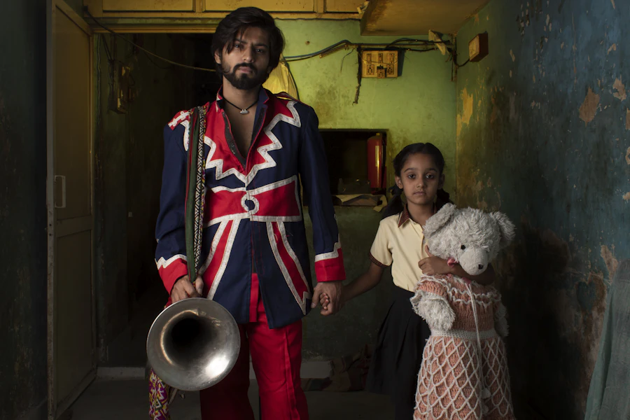 A man in a colorful Union Jack-style outfit stands beside a young girl holding a teddy bear in a worn interior room. Warm light falls across their faces against a dark doorway and peeling blue wall.