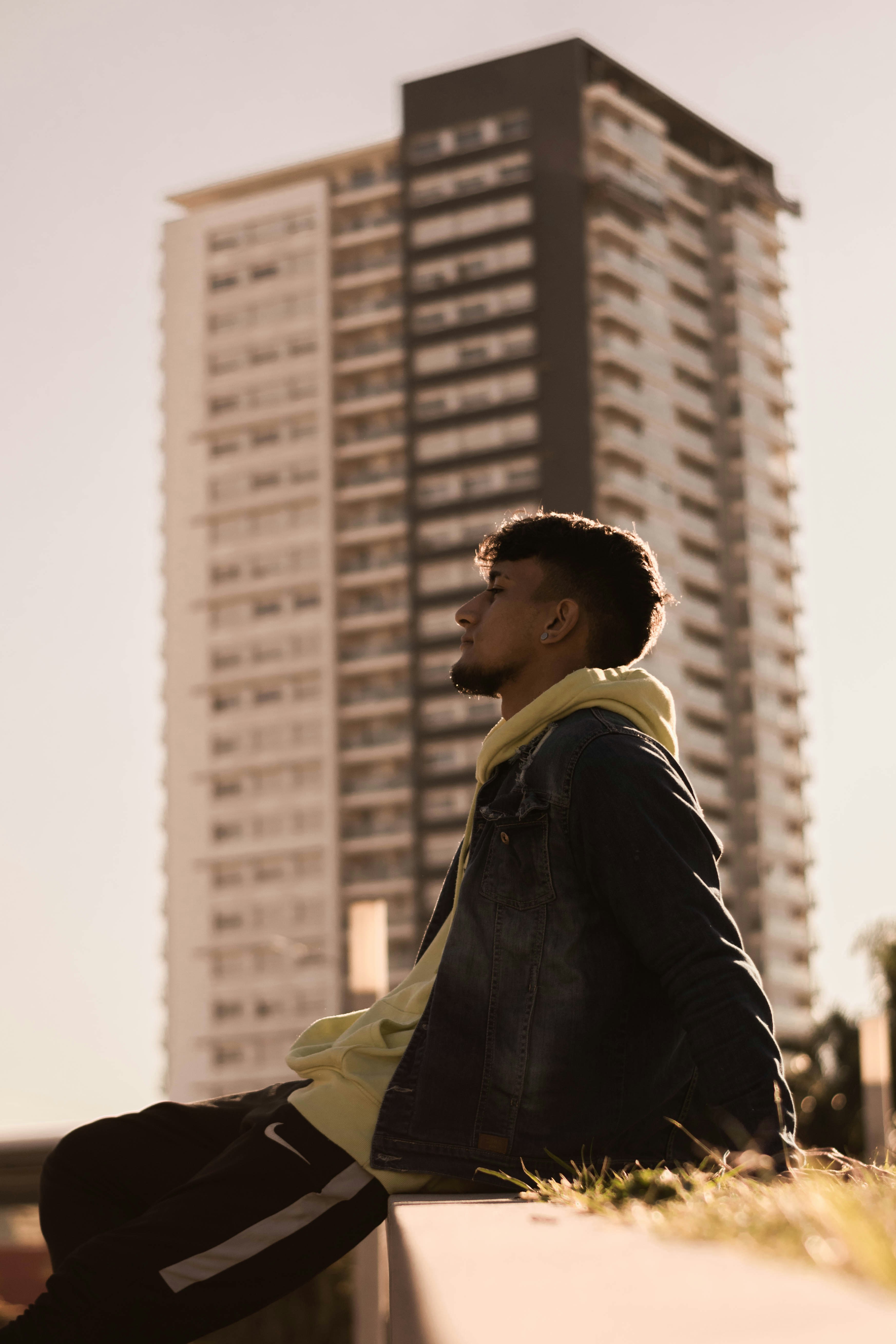 Young man in a denim jacket and hoodie sits thoughtfully against a backdrop of a modern high-rise building. The warm light accentuates the scene.