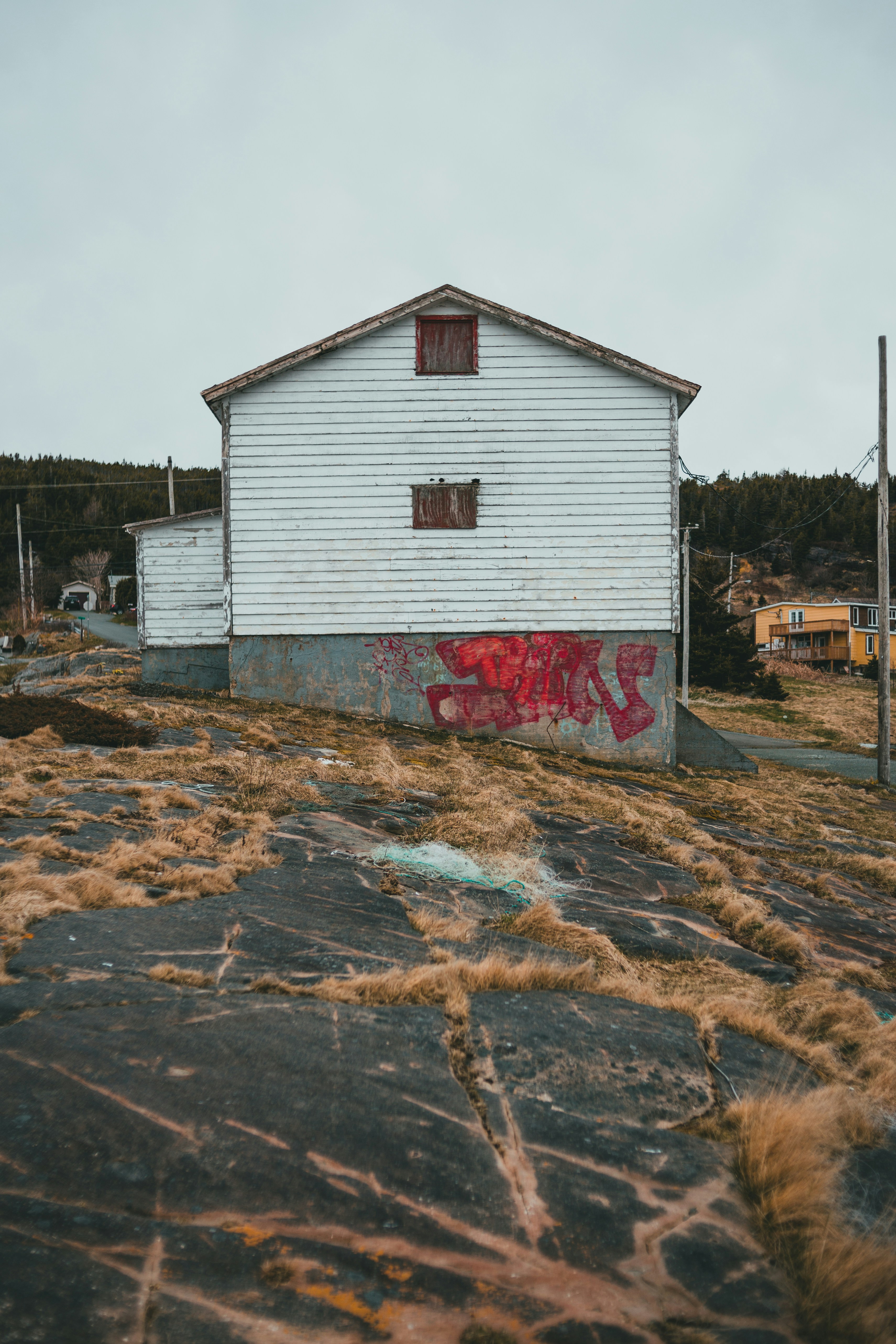 A weathered white house with boarded windows and vibrant graffiti stands on rocky terrain, surrounded by sparse grass and distant homes. The scene evokes a sense of nostalgia and decay.