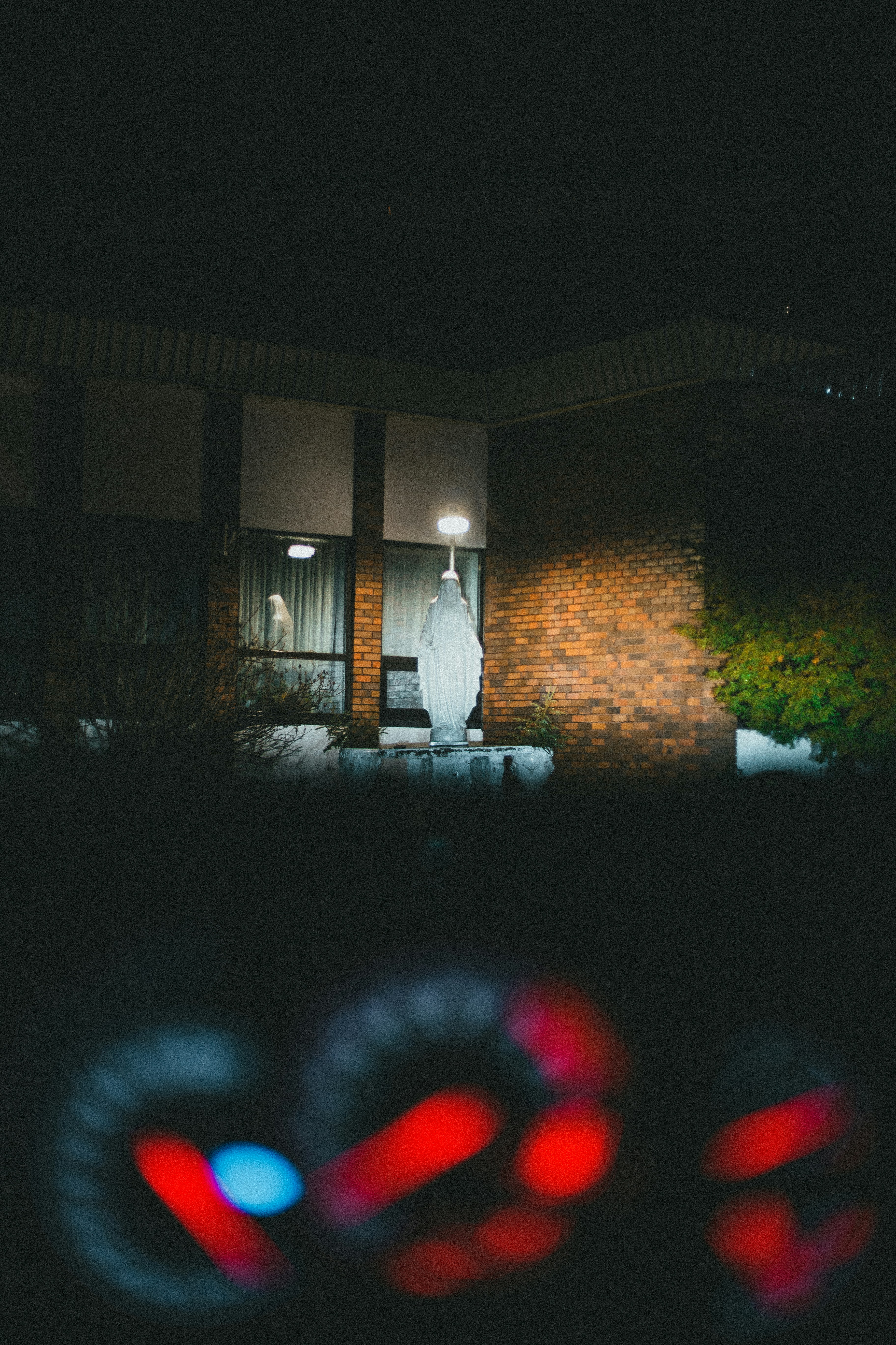 Nighttime vigil by the lit doorway: a ghostly figure in a blue dress on a brick facade