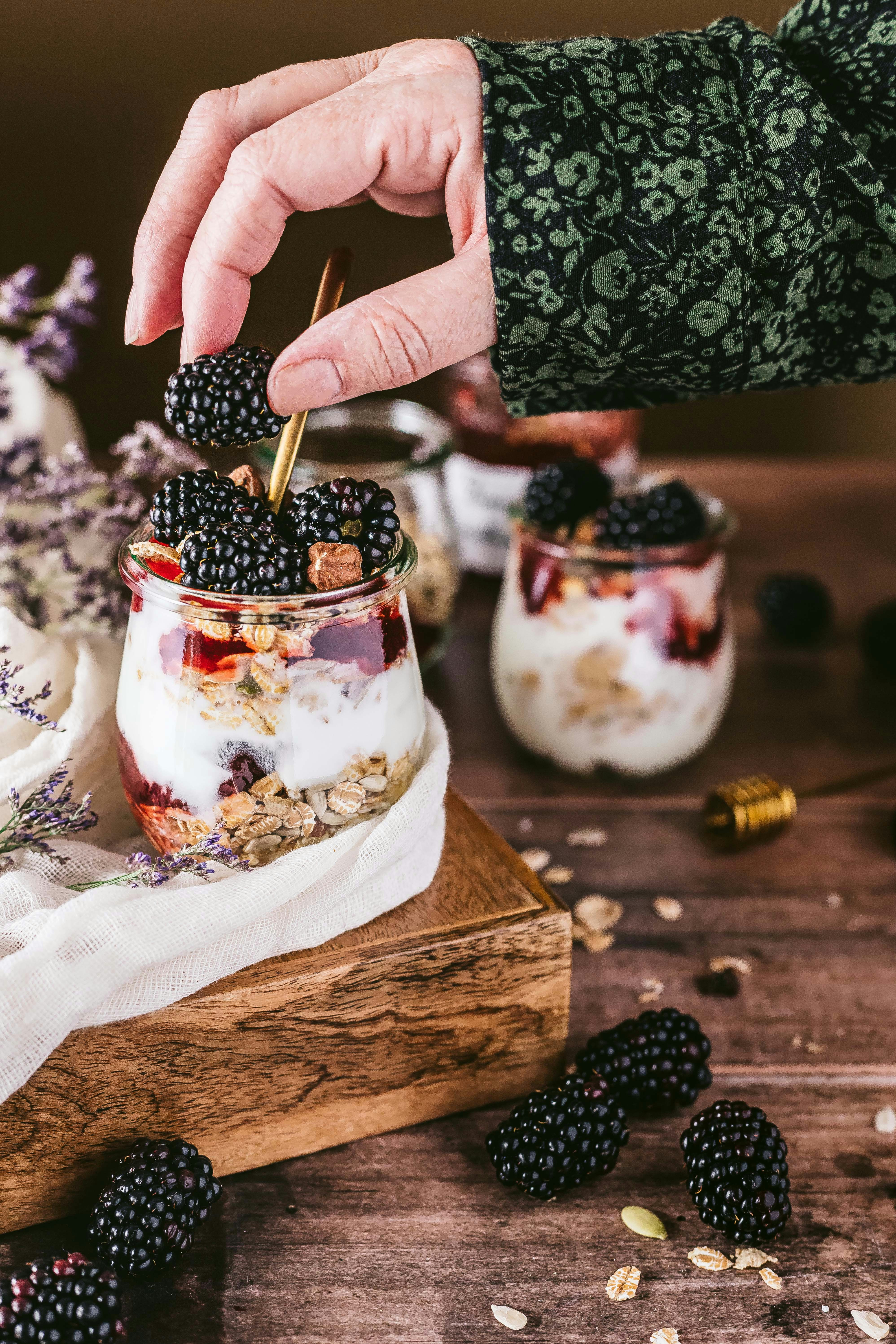 Hand placing fresh blackberries atop layered yogurt and granola in a glass jar, surrounded by lavender and scattered berries.