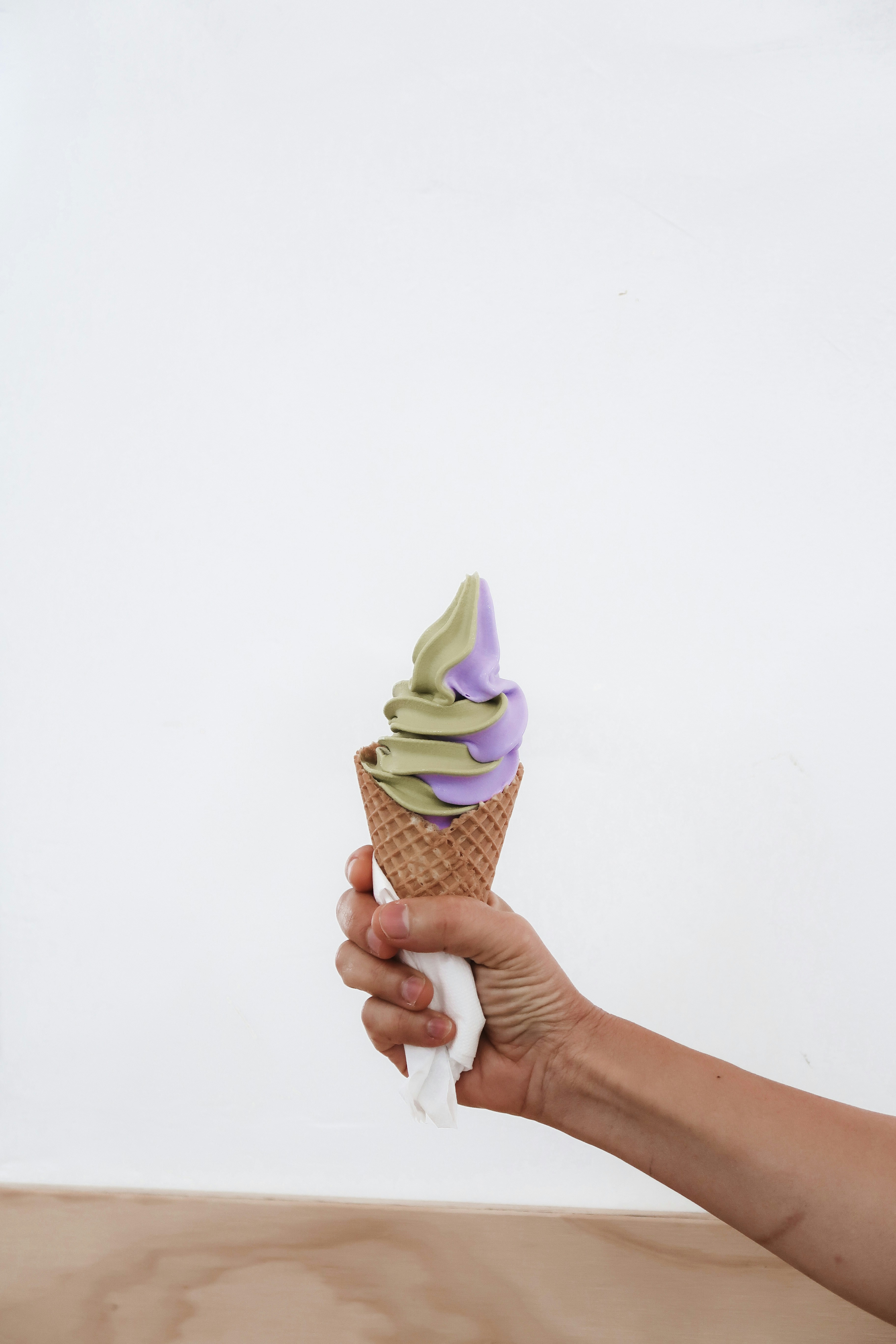 A hand grips a waffle cone topped with lavender and olive pastel swirls against a plain white backdrop.