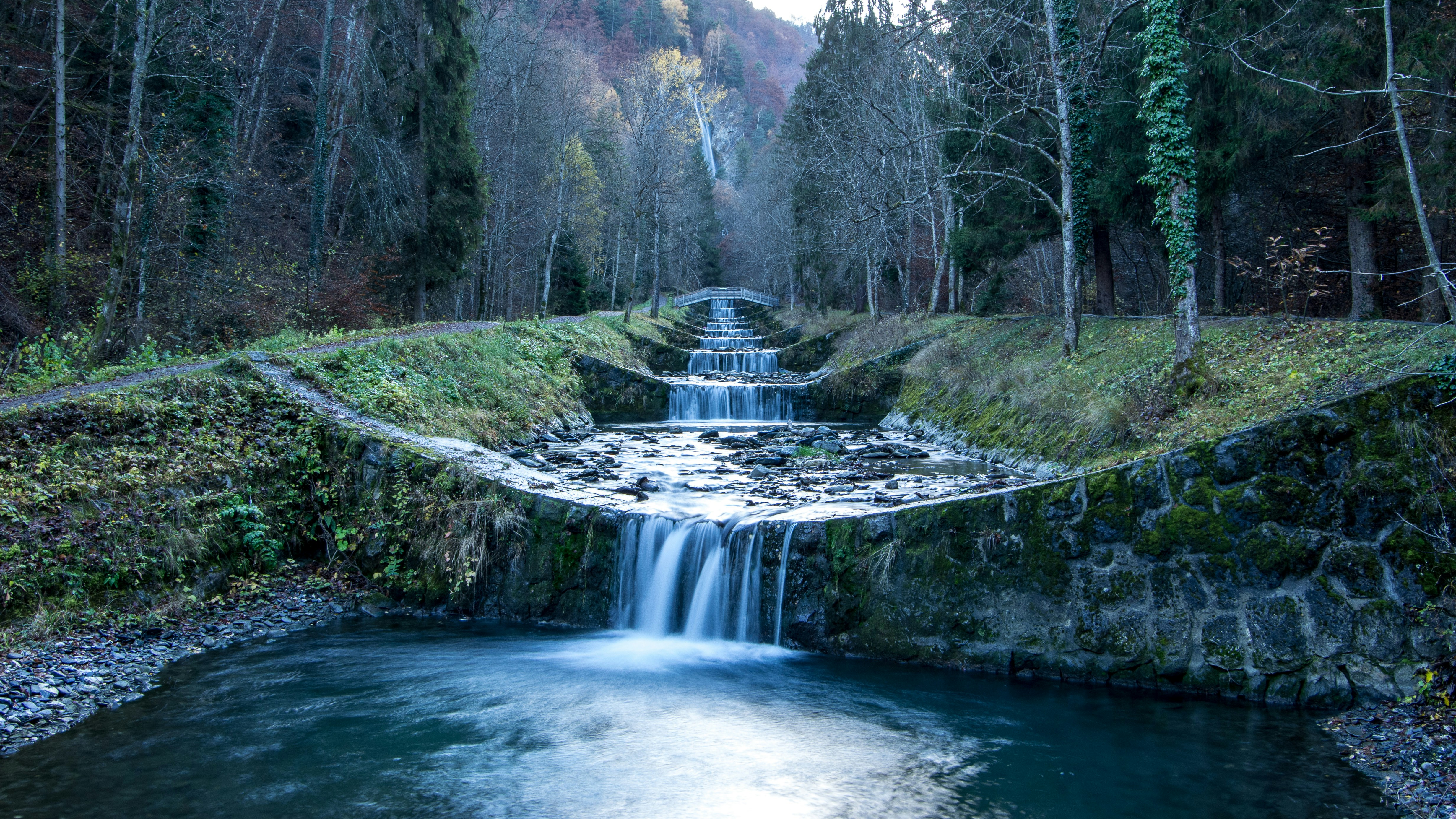 Gentle waterfalls cascading over stone steps, surrounded by lush greenery and autumn foliage. A tranquil stream reflects the serene landscape.