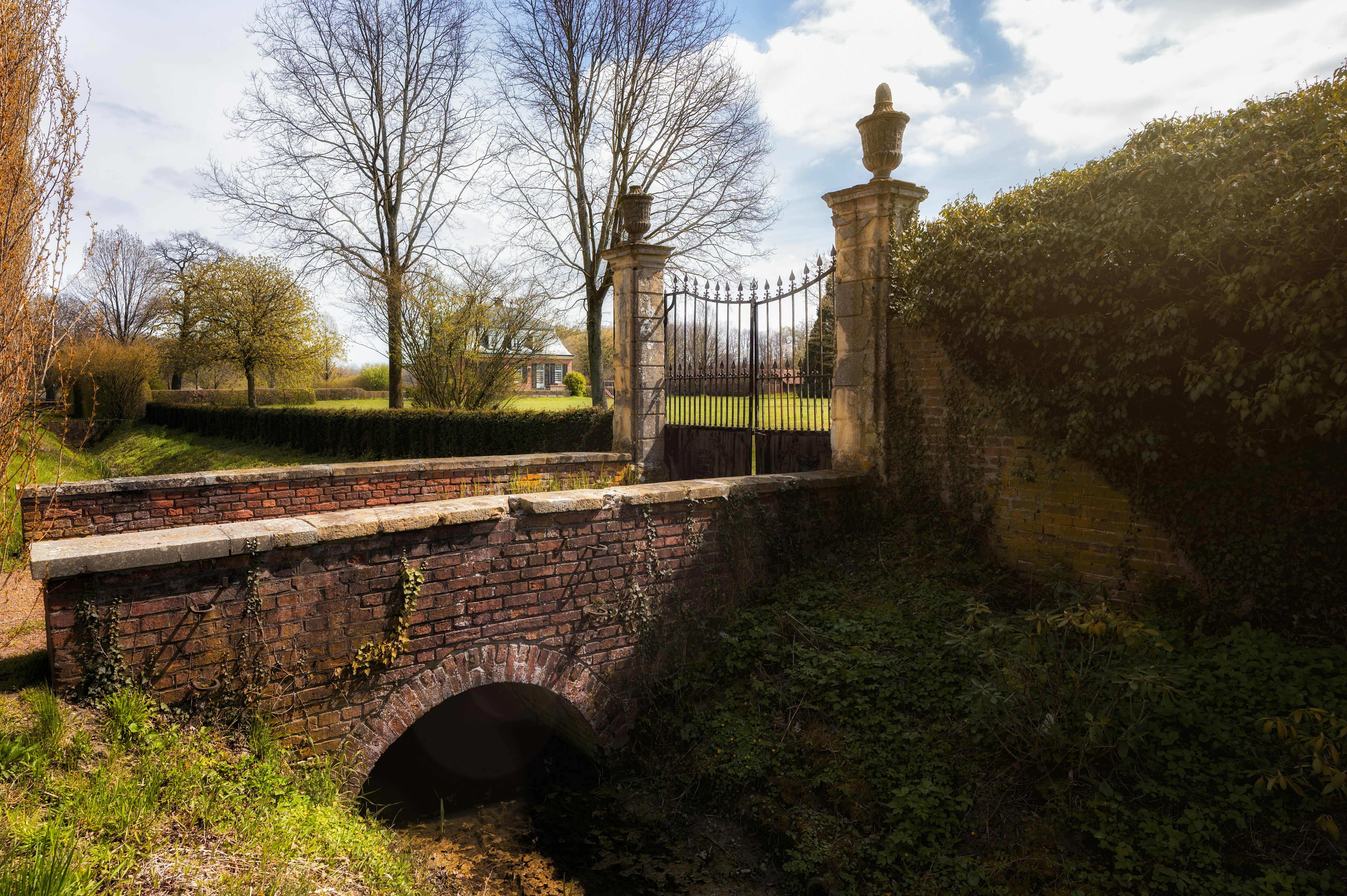 Brick bridge arching over a small stream, flanked by ornate gateposts and lush greenery, leading to a distant estate. The scene evokes a sense of history and tranquility.