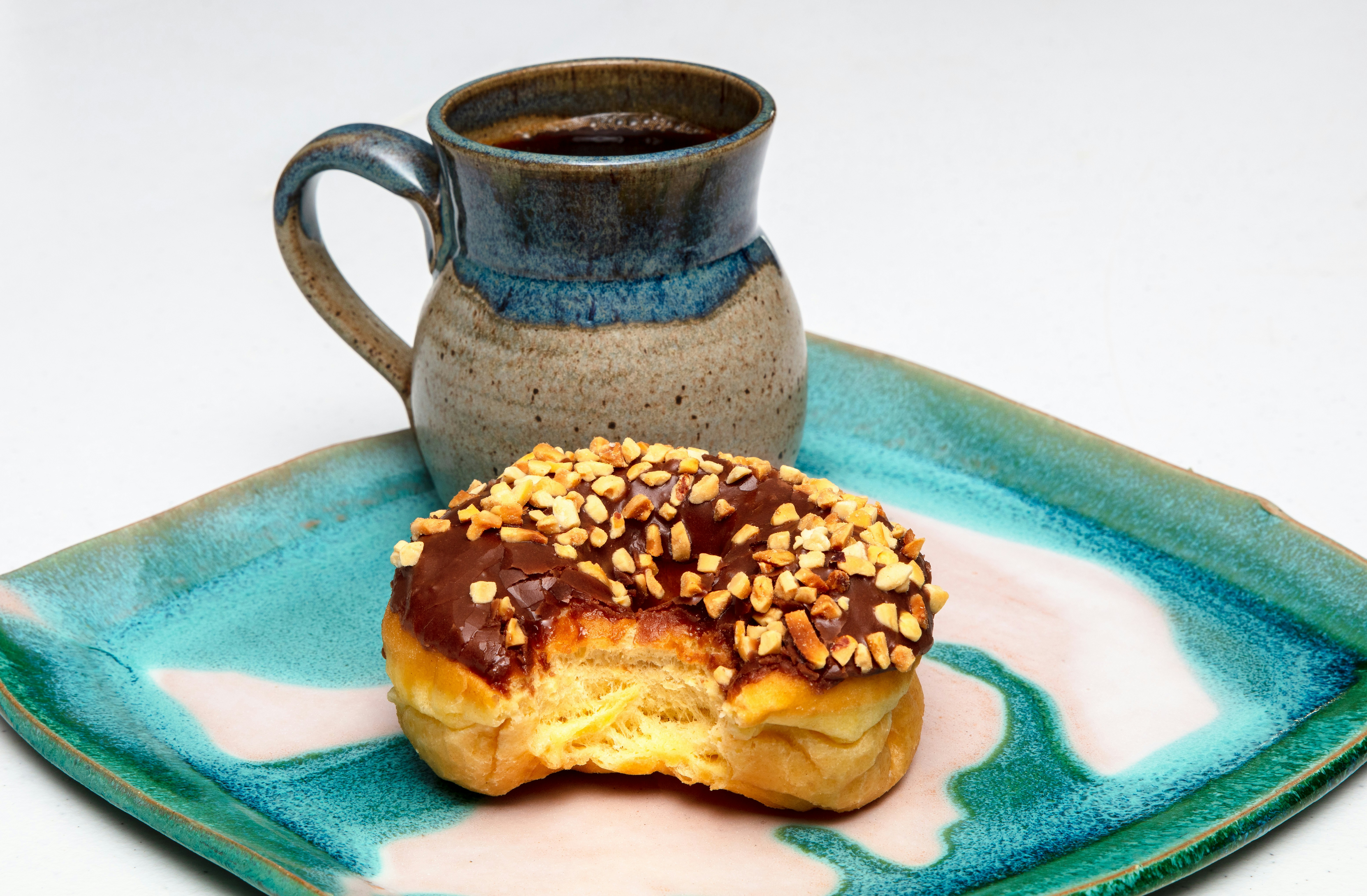 Chocolate-covered donut topped with crushed nuts beside a rustic ceramic mug filled with coffee on a colorful plate.