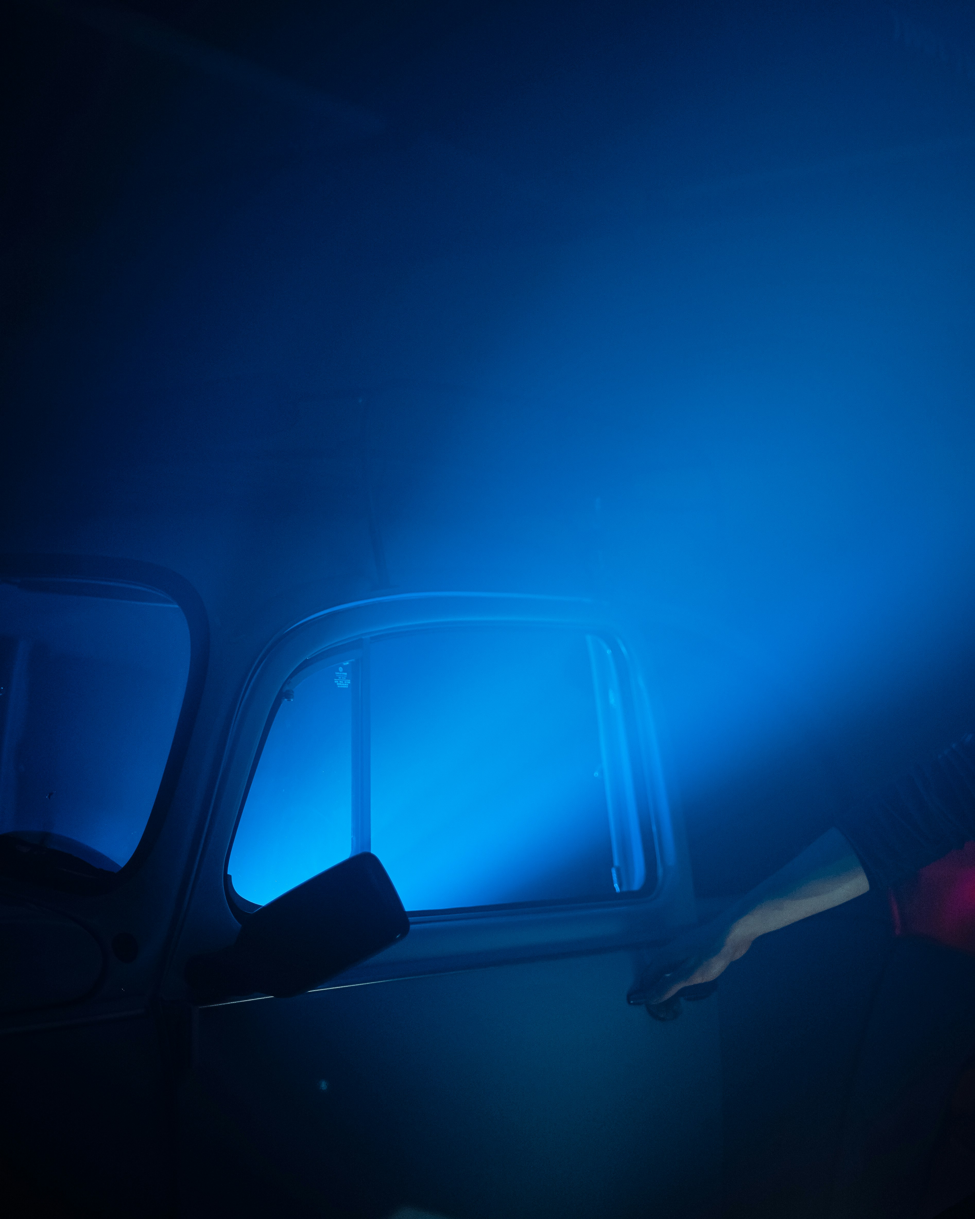 A hand reaches towards an old car door, illuminated by a beam of blue light cutting through the darkness. The scene evokes a sense of nostalgia and mystery.