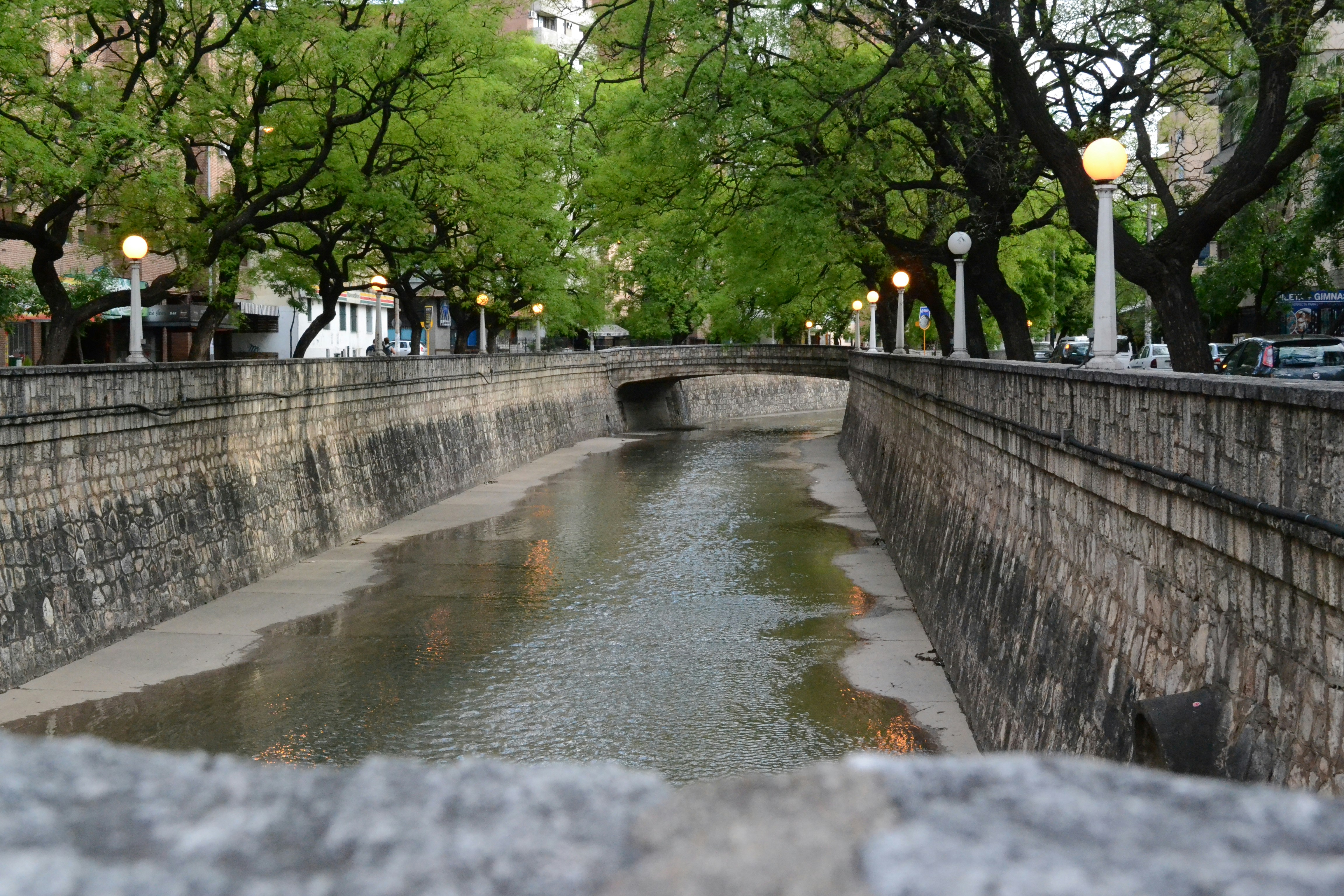 Narrow stone-lined canal flanked by lush green trees and vintage street lamps in an urban setting.