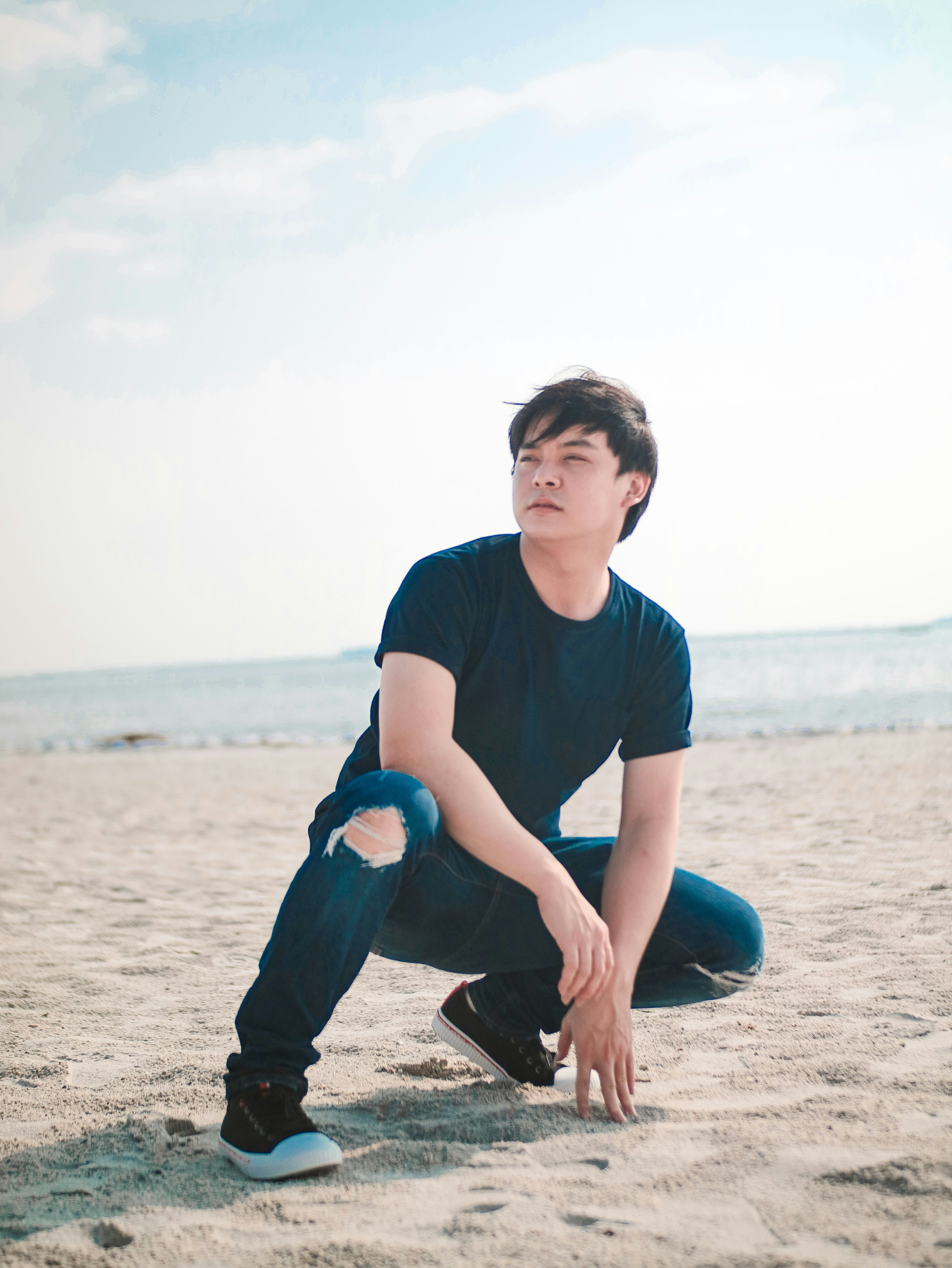 Young man crouching on sandy beach, gazing thoughtfully towards the horizon. Soft waves and clear sky create a serene backdrop.