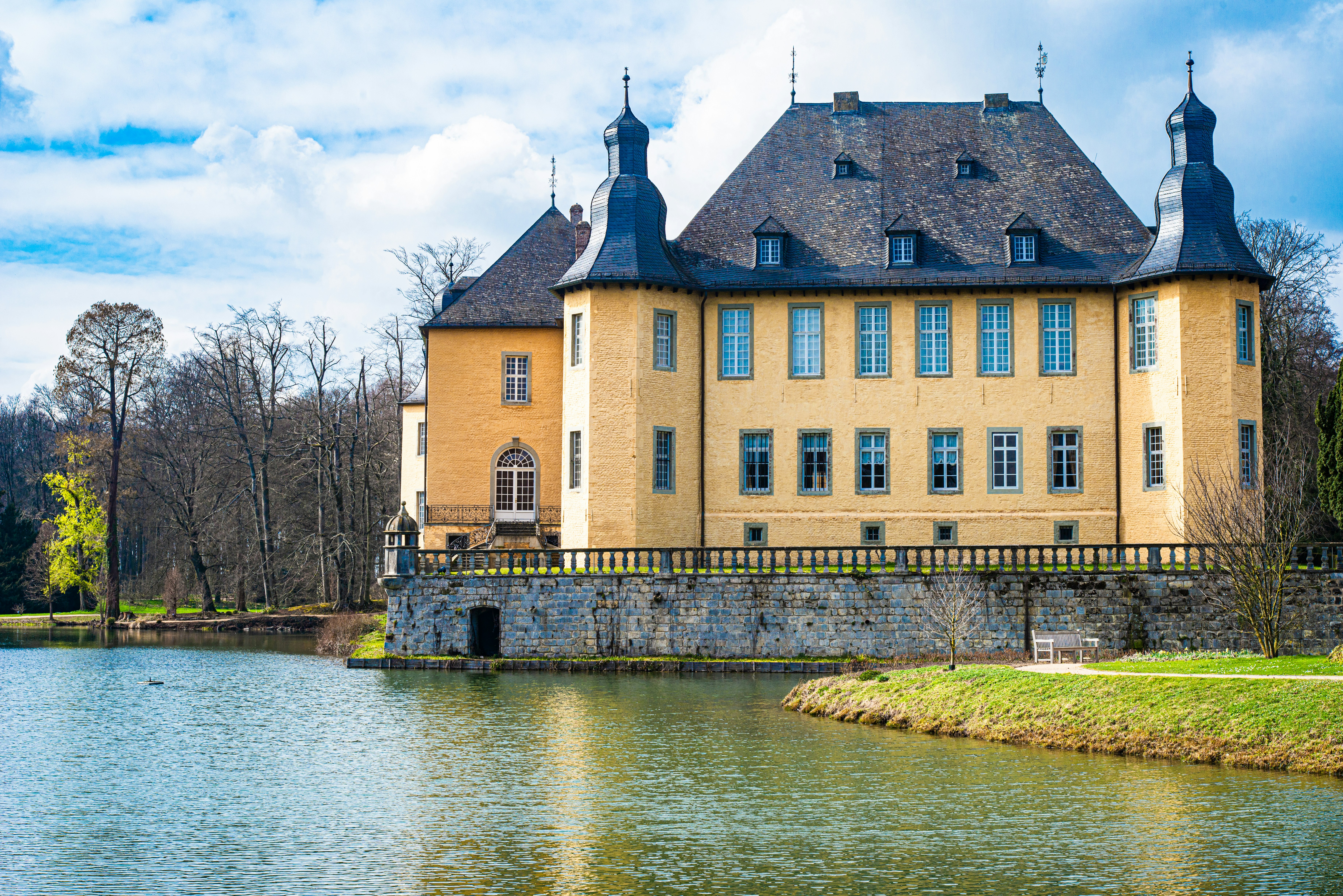 Yellow castle with turrets reflected in a tranquil lake, surrounded by bare trees and a cloudy sky.