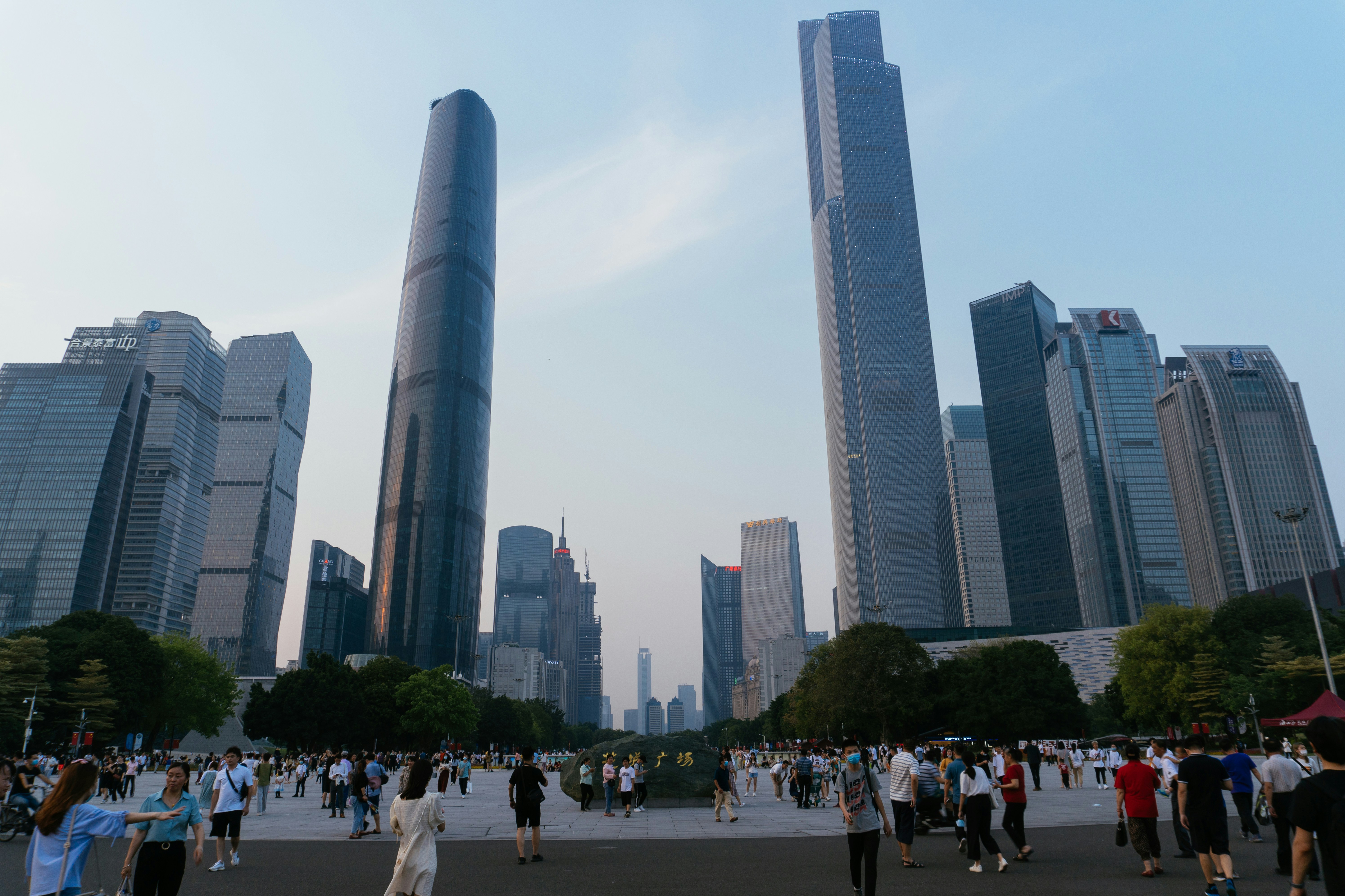 People gather in a bustling city square framed by towering skyscrapers against a clear sky.