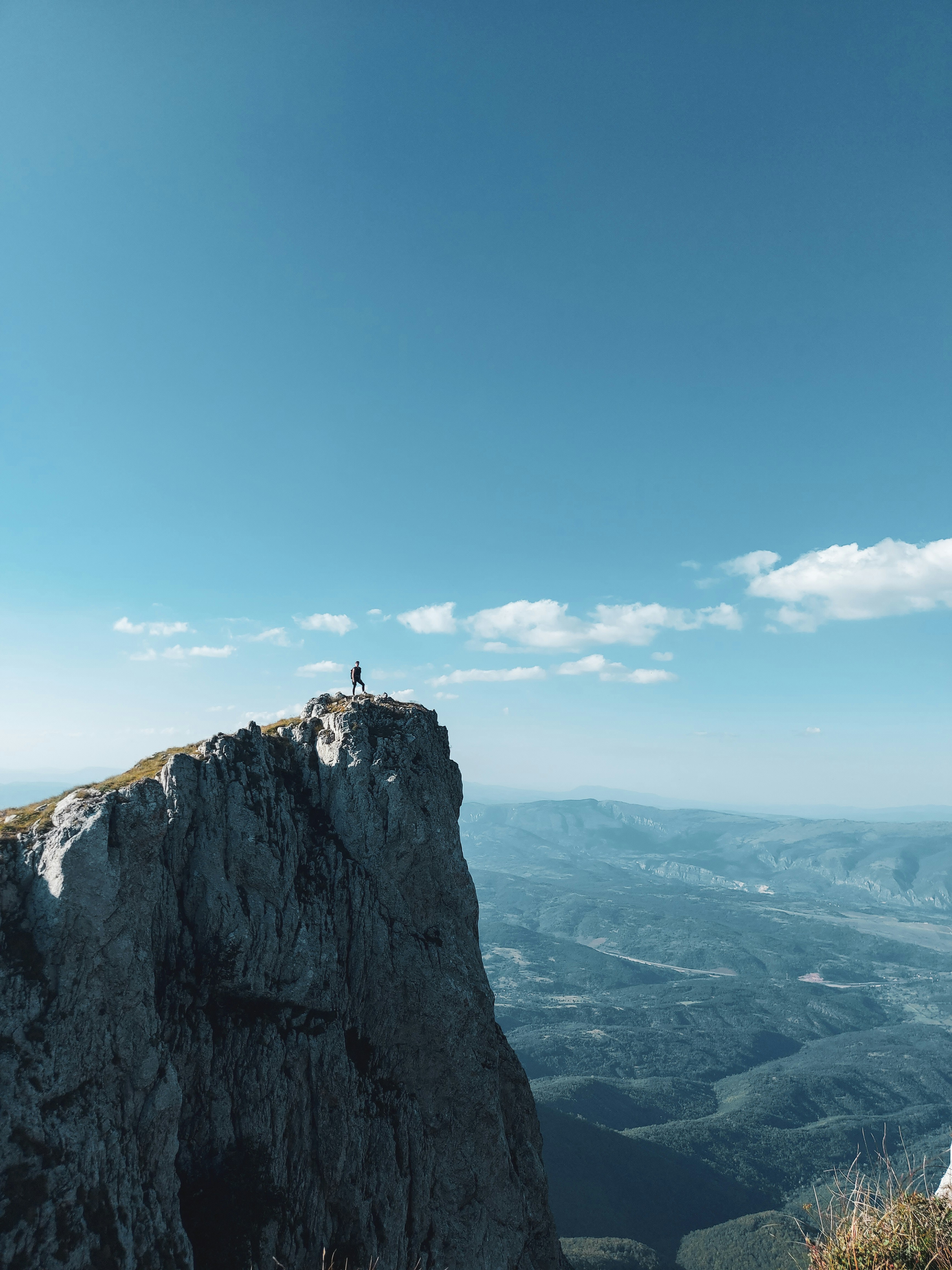 A lone figure stands atop a rugged mountain peak, overlooking a vast valley under a clear blue sky. The scene captures the essence of adventure and the beauty of nature.