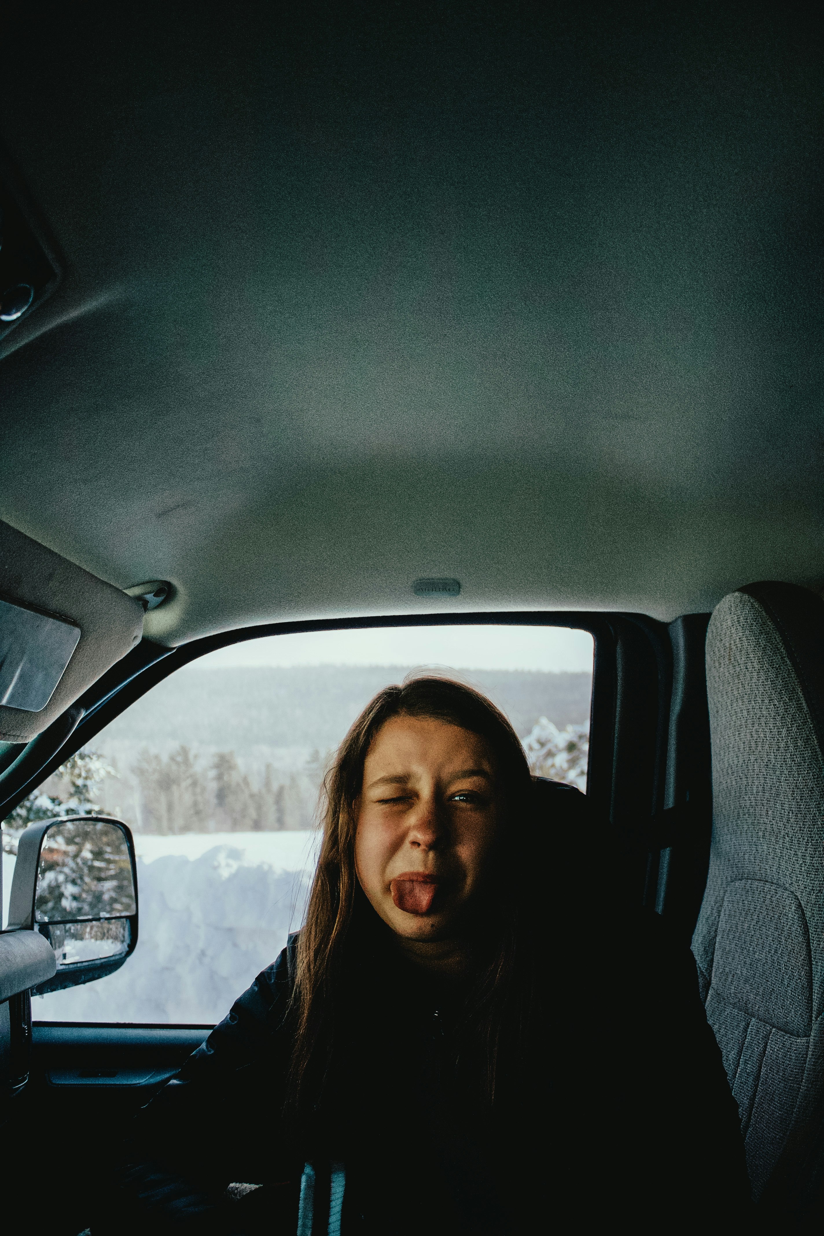 A young woman playfully sticking her tongue out inside a vehicle, with a scenic snowy landscape visible through the window.