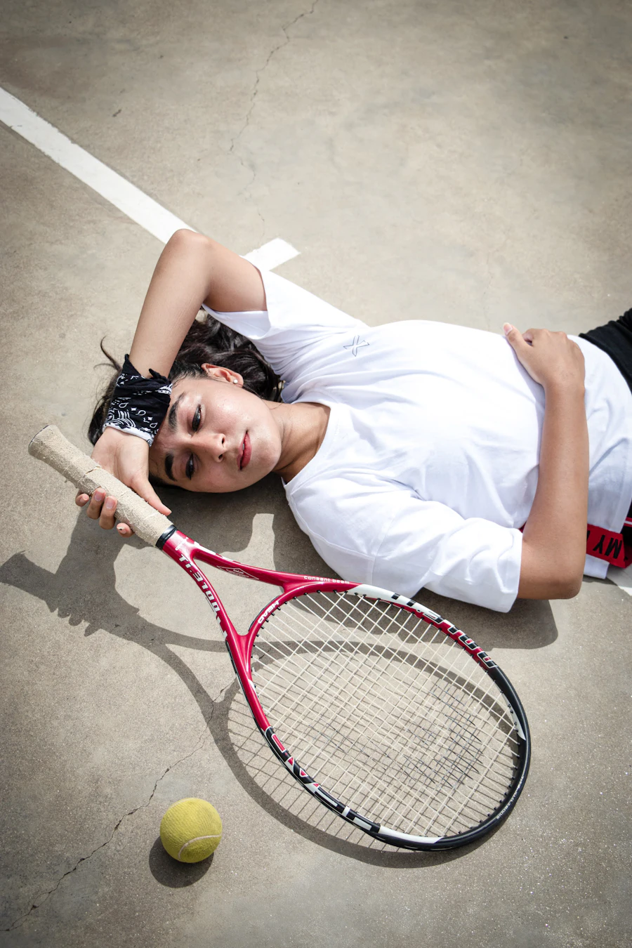 Young woman lying on a concrete tennis court holding a red racket with a tennis ball close by.