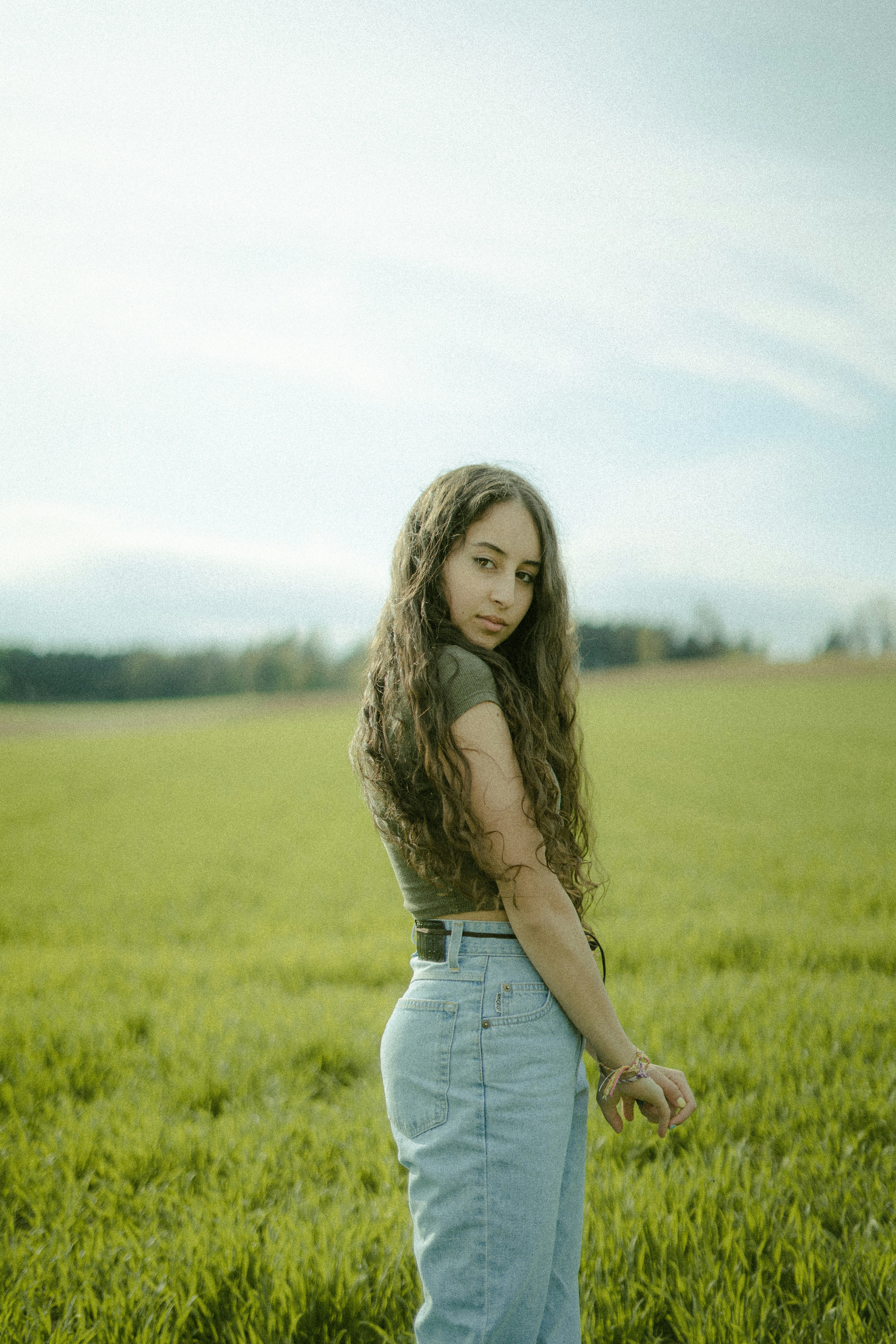 Young woman with long curly hair stands in a vibrant green field, gazing over her shoulder against a soft sky backdrop.