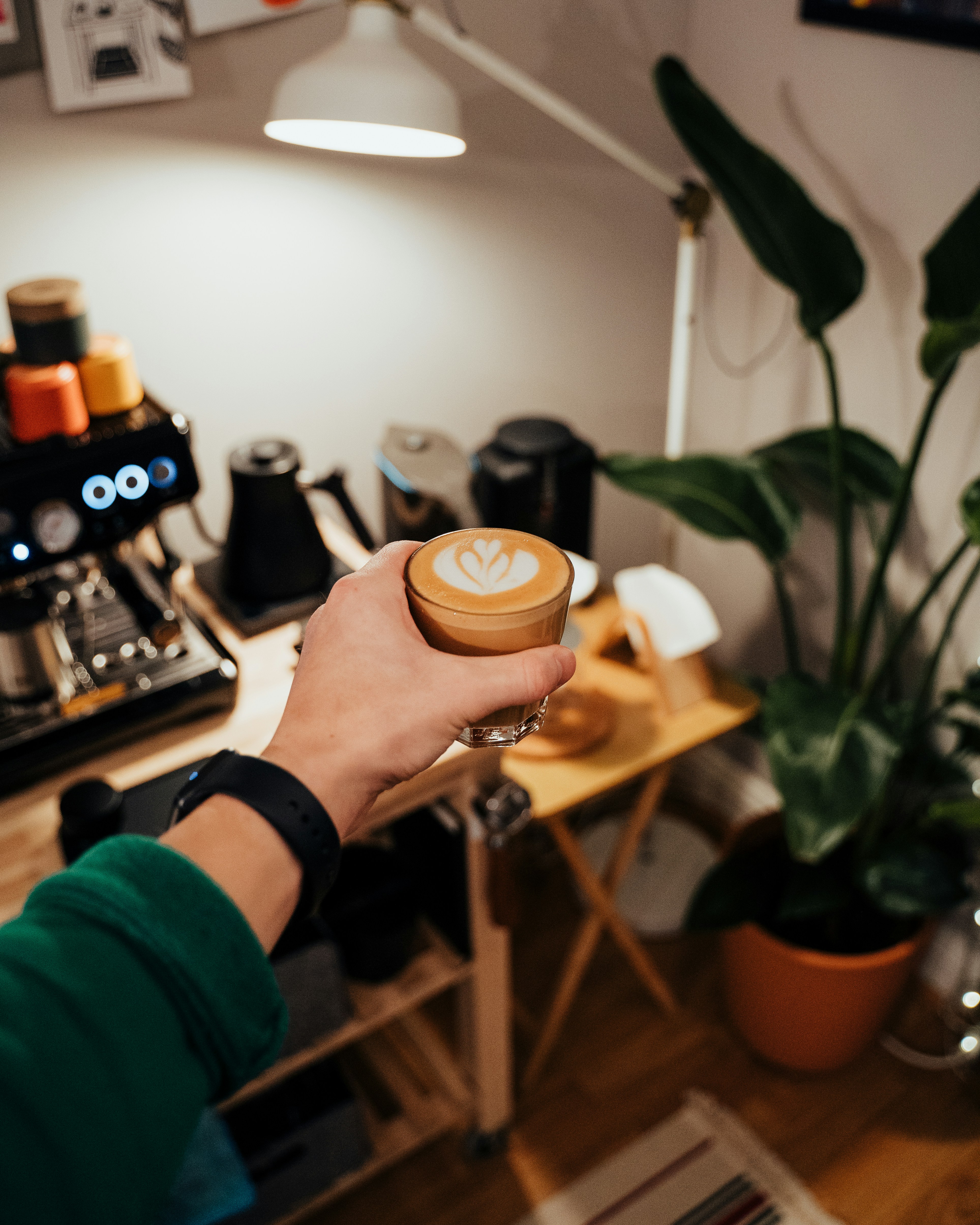 Hand holding a latte with intricate foam art against a backdrop of coffee equipment and greenery. 