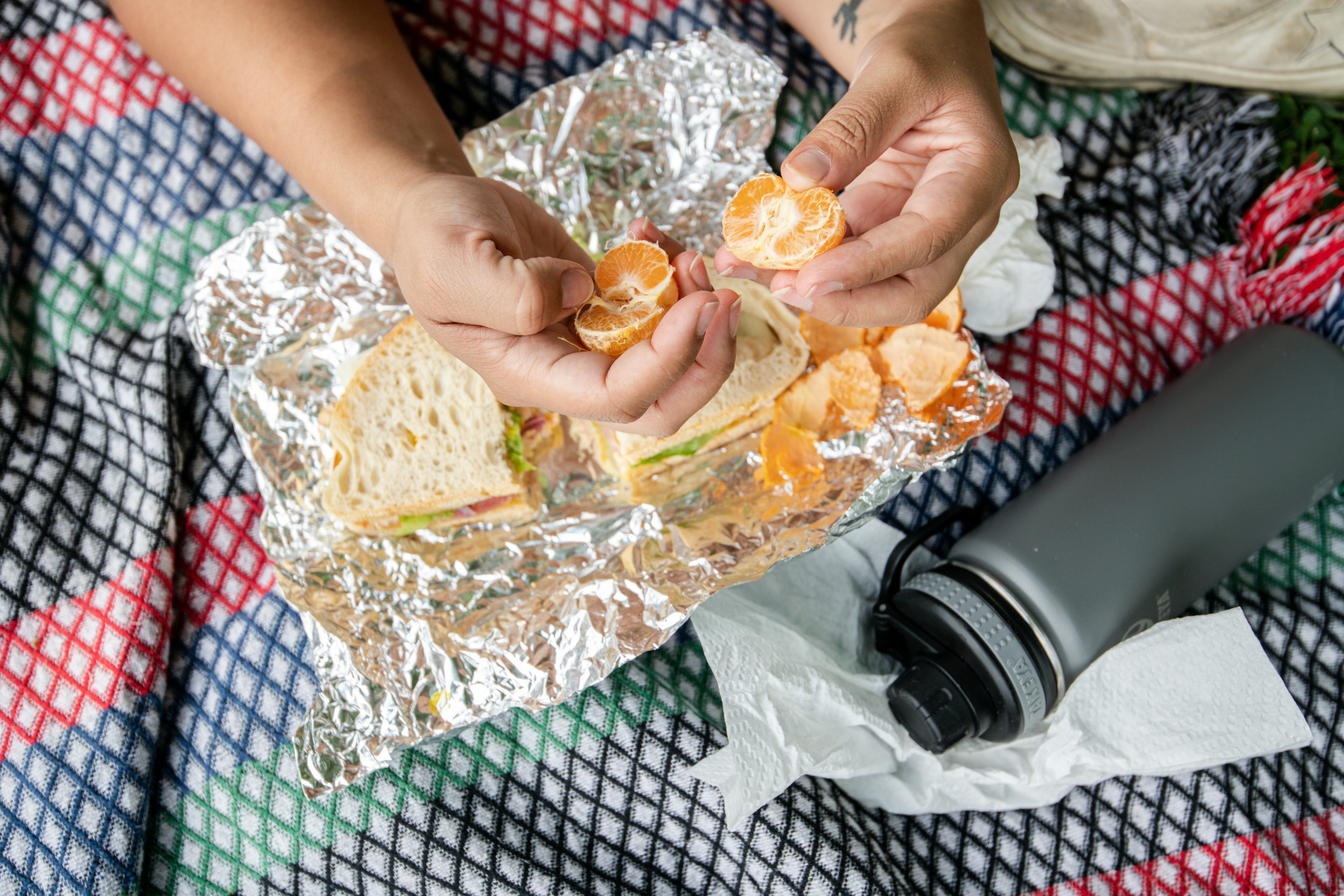 Hands delicately peel citrus while a sandwich rests in aluminum foil on a checkered blanket. A water bottle is nearby, suggesting a relaxed outdoor setting.