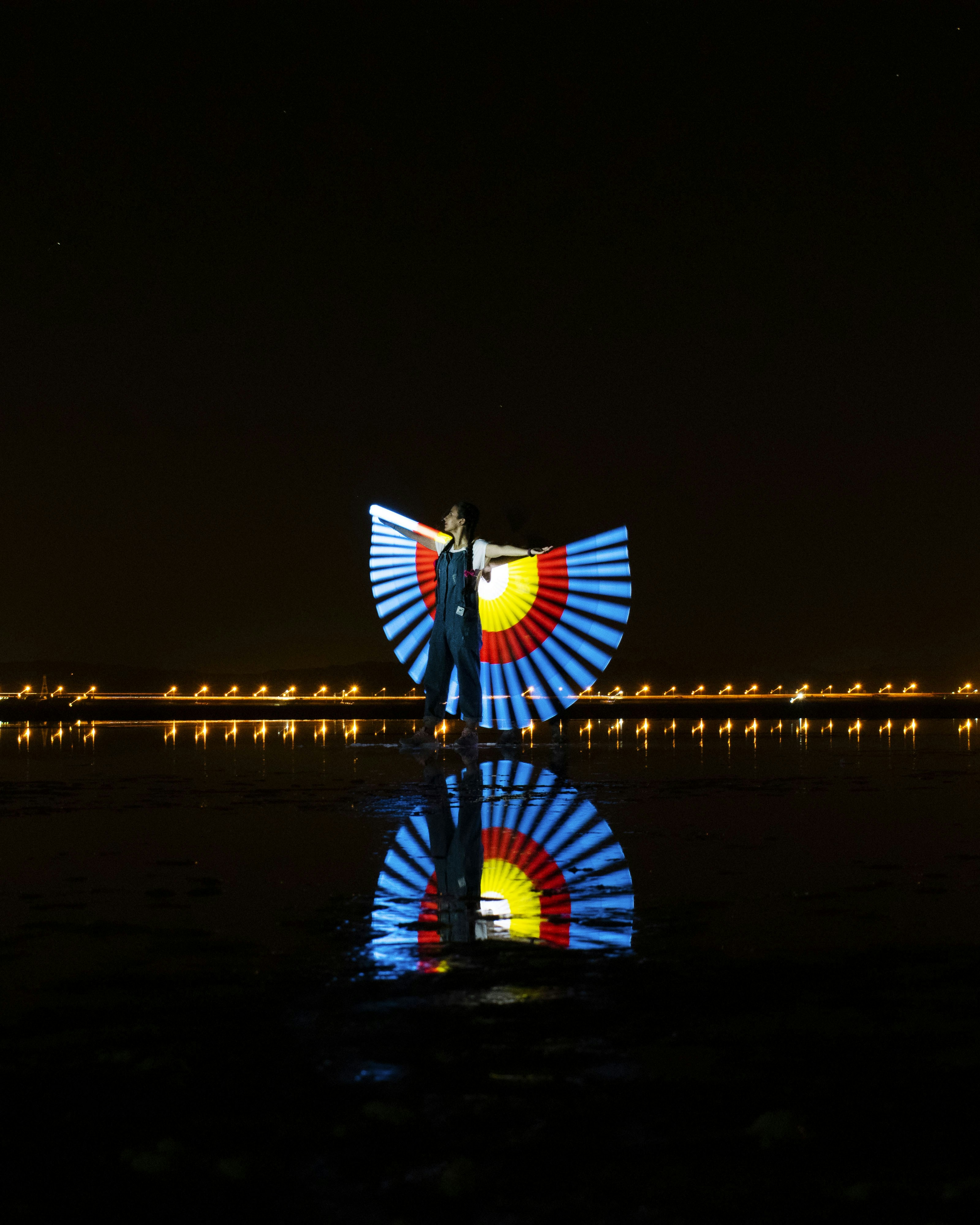 A performer showcases vibrant illuminated fans against a dark backdrop, reflected on a calm water surface.
