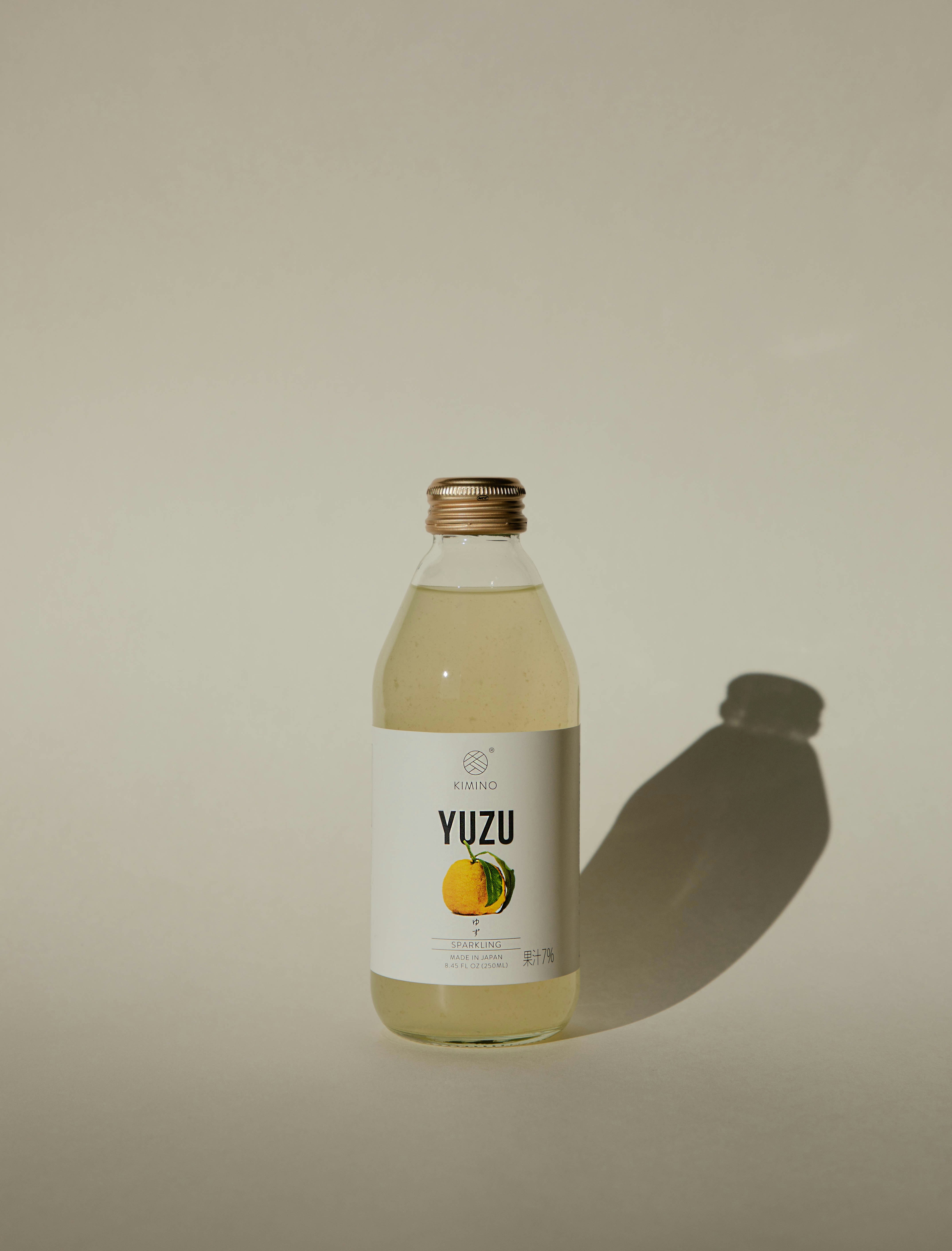 A bottle of yuzu beverage stands against a soft, neutral backdrop, showcasing its label and refreshing contents. The subtle shadow adds depth to the composition.