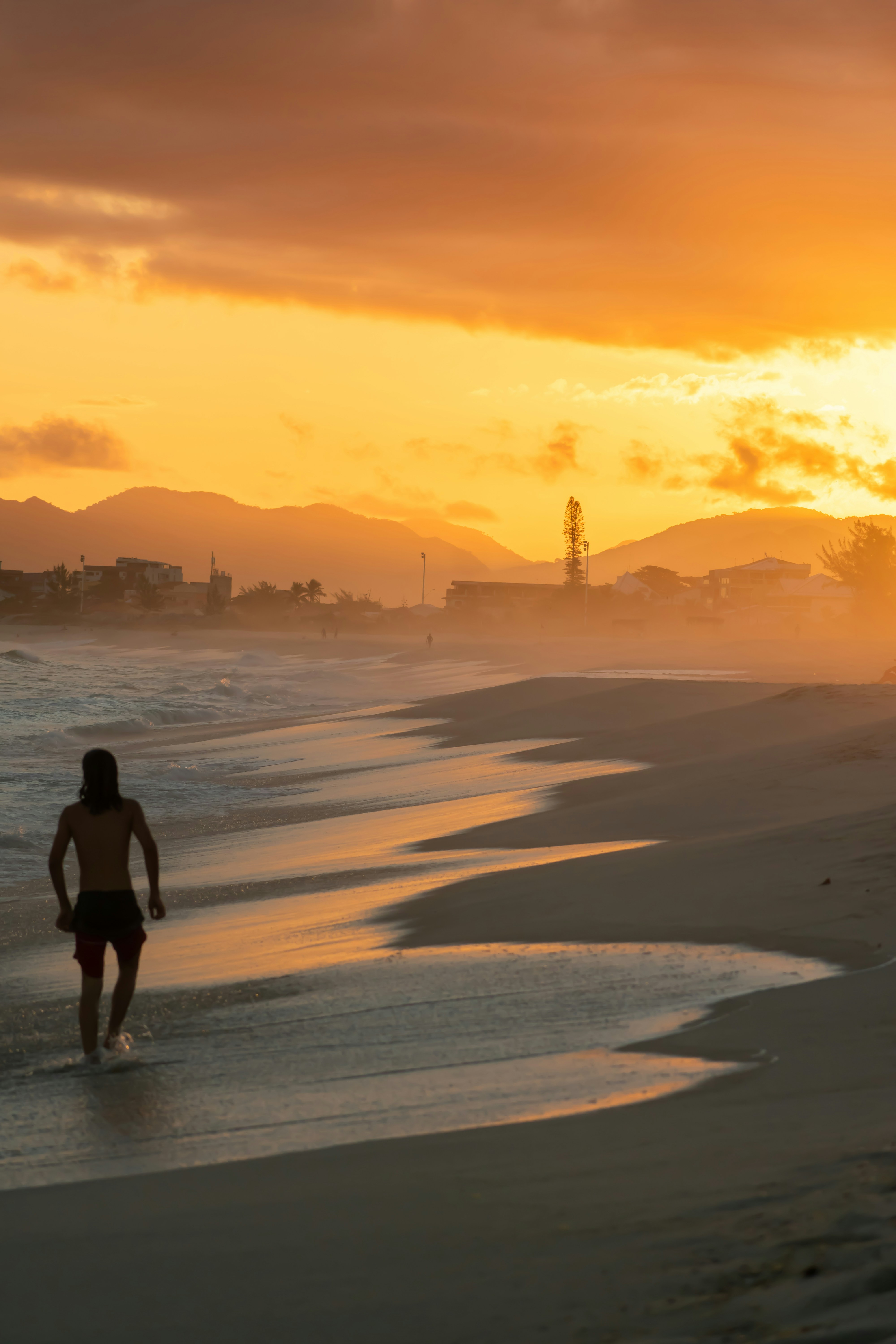 Silhouette of a person walking along a tranquil beach at sunset, with waves gently lapping at the shore. The sky is painted in warm hues of orange and gold.