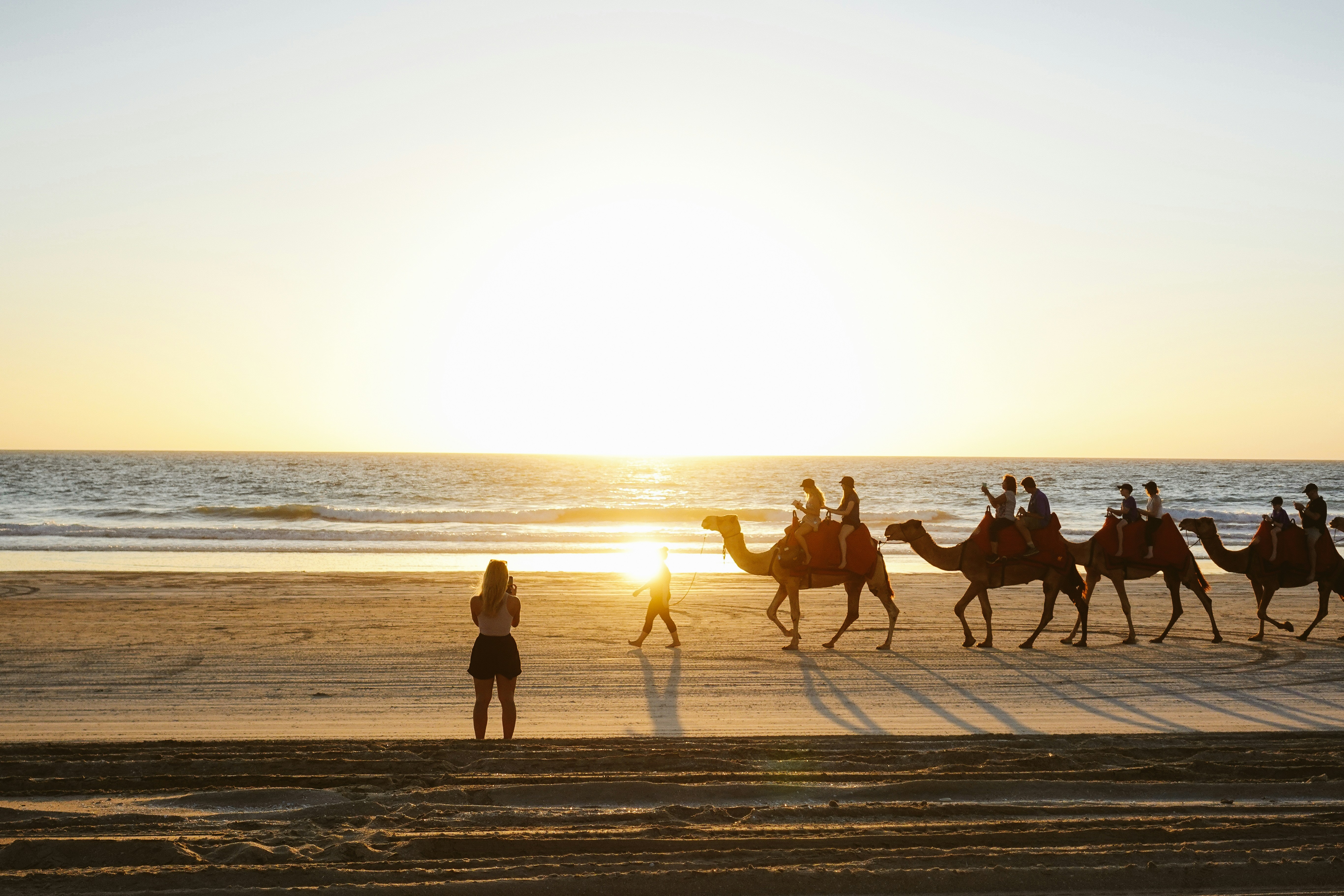 Silhouetted camels walk along the beach at sunset, with a lone figure capturing the moment. The warm glow of the setting sun casts long shadows on the sand.