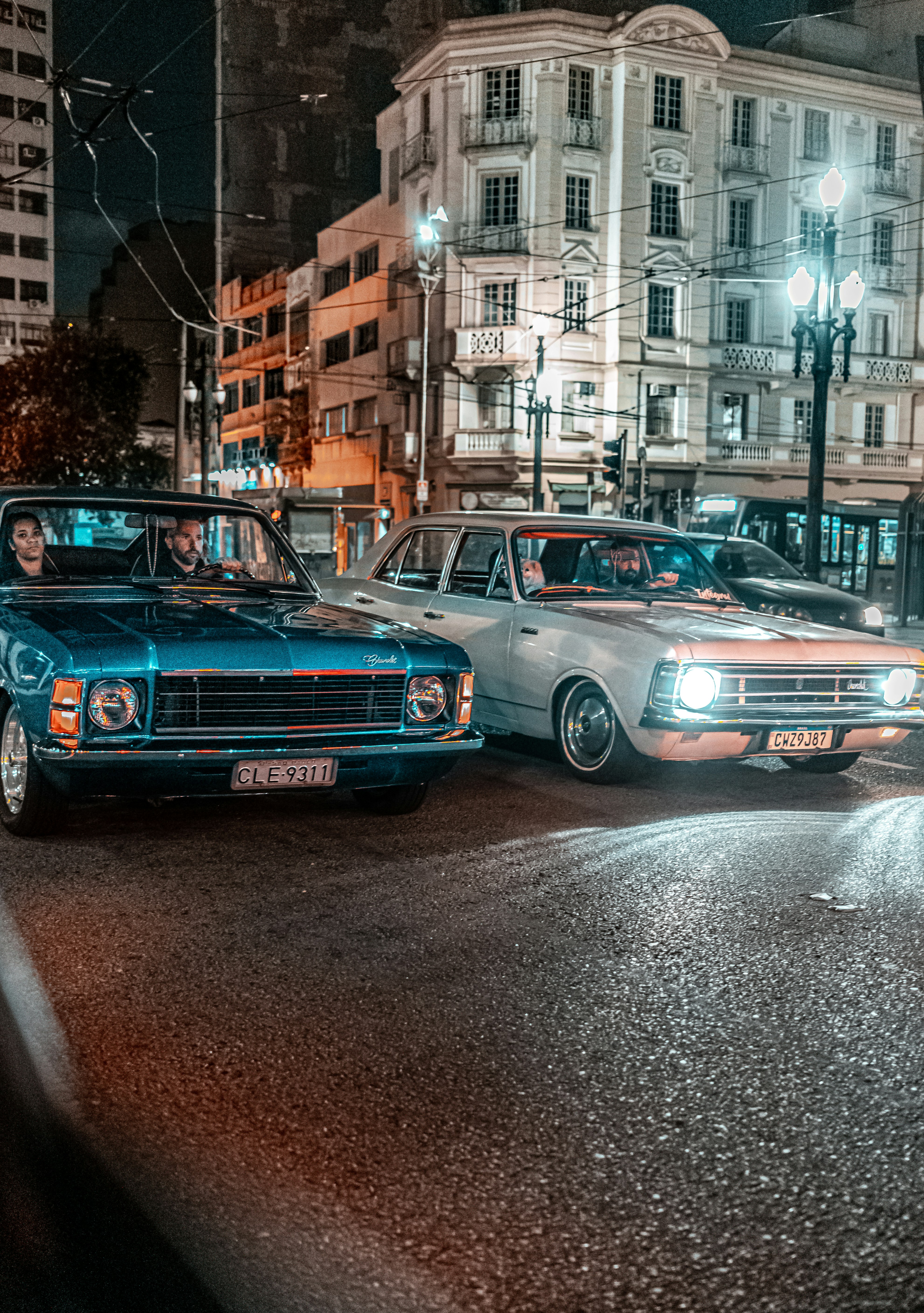 Two classic cars, a blue sedan and a white coupe, parked at a city intersection illuminated by streetlights at night.