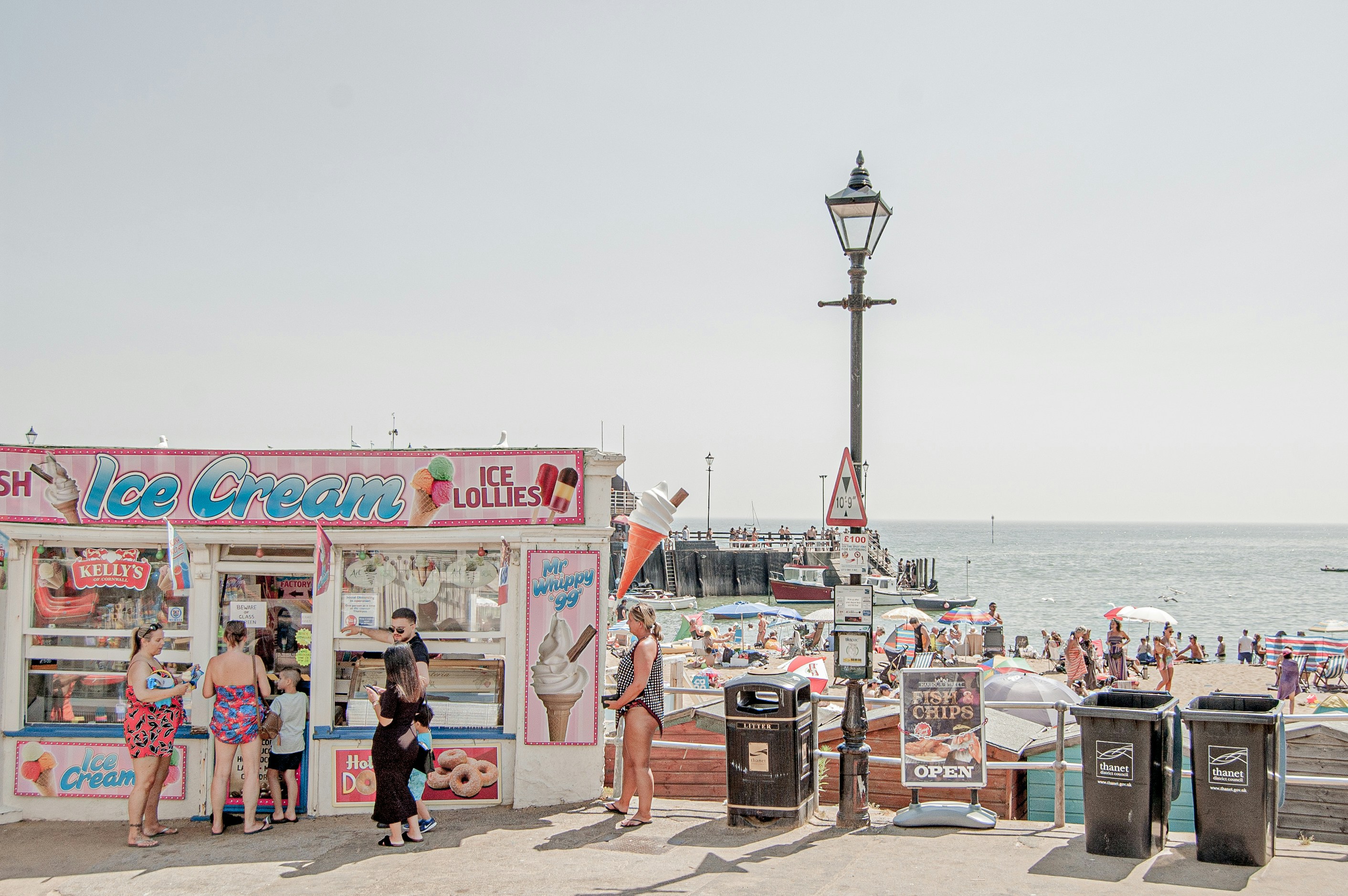 Ice cream shop beside a busy promenade with beachgoers and a distant pier.